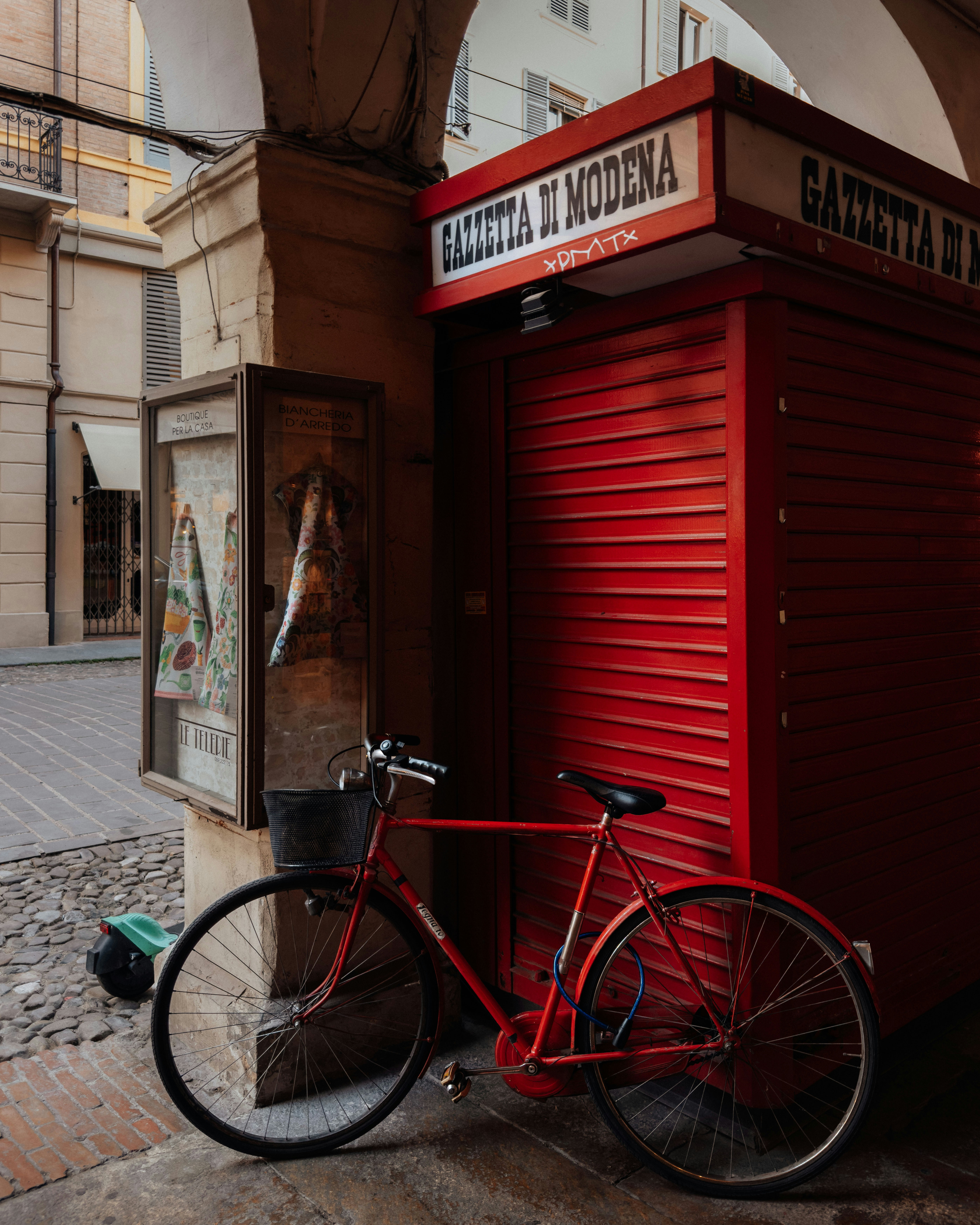 Red bicycle leans against a bold red shutter beneath a stone arch on a quiet Italian street. The scene emphasizes urban character and architectural detail.