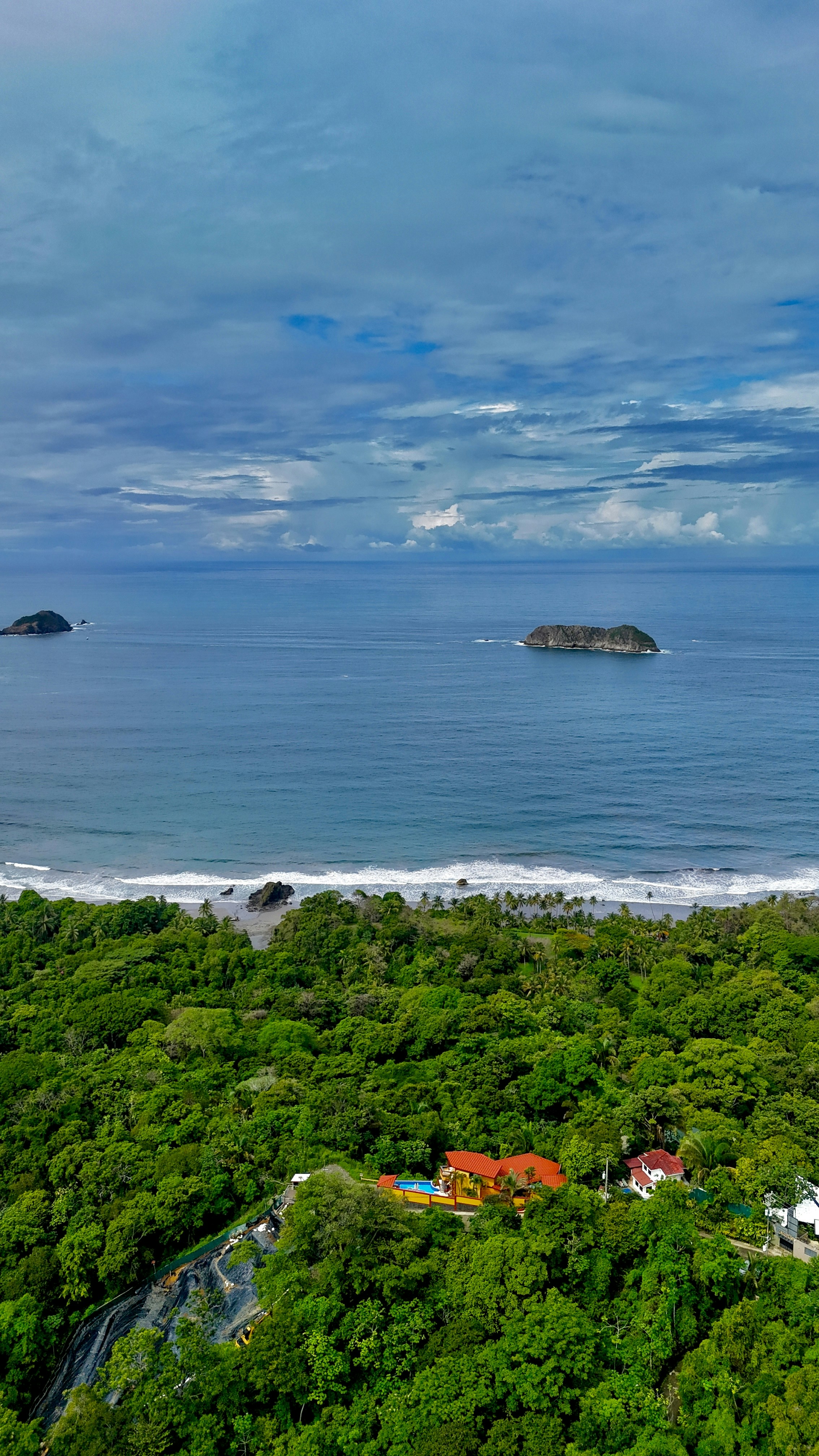 Aerial photograph of a dense tropical forest meeting a tranquil blue sea, with two small islands on the horizon and red-roofed buildings at the forest edge.