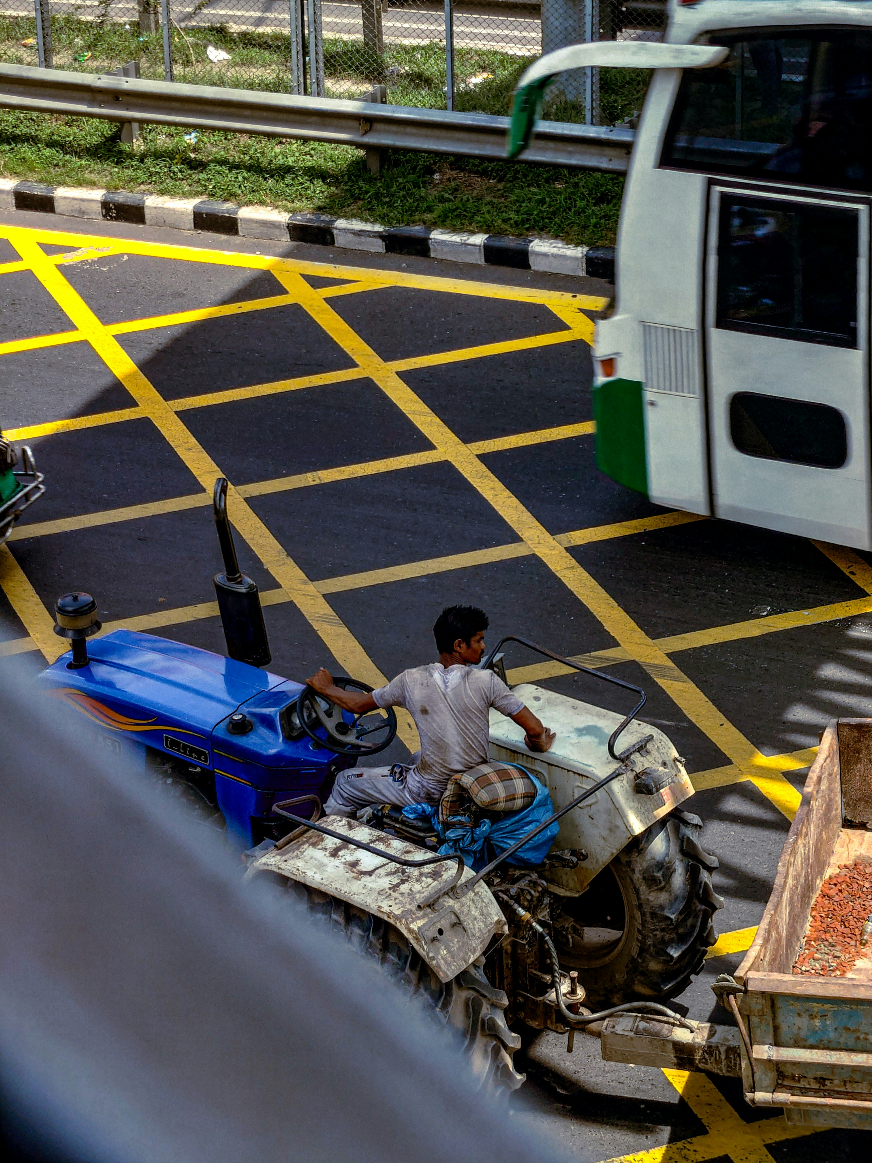 A street-scene photograph of a man driving a blue tractor along bold yellow crosshatched markings, with a white bus nearby in an urban setting.