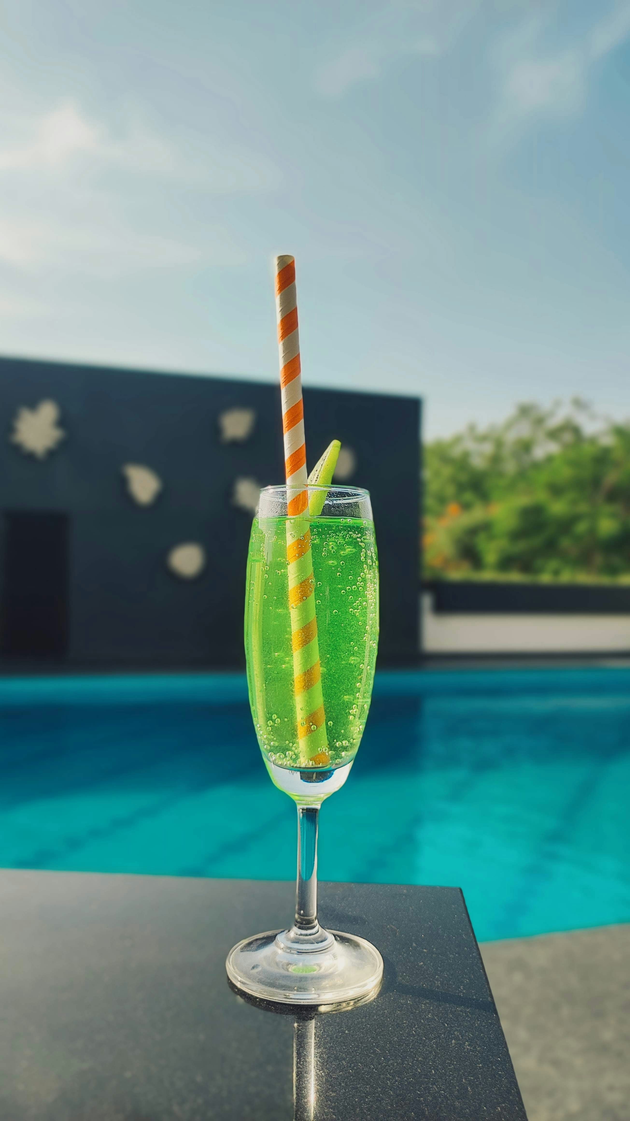 Vibrant green drink in a tall glass with a striped straw, set against a sparkling blue pool and a minimalist backdrop.