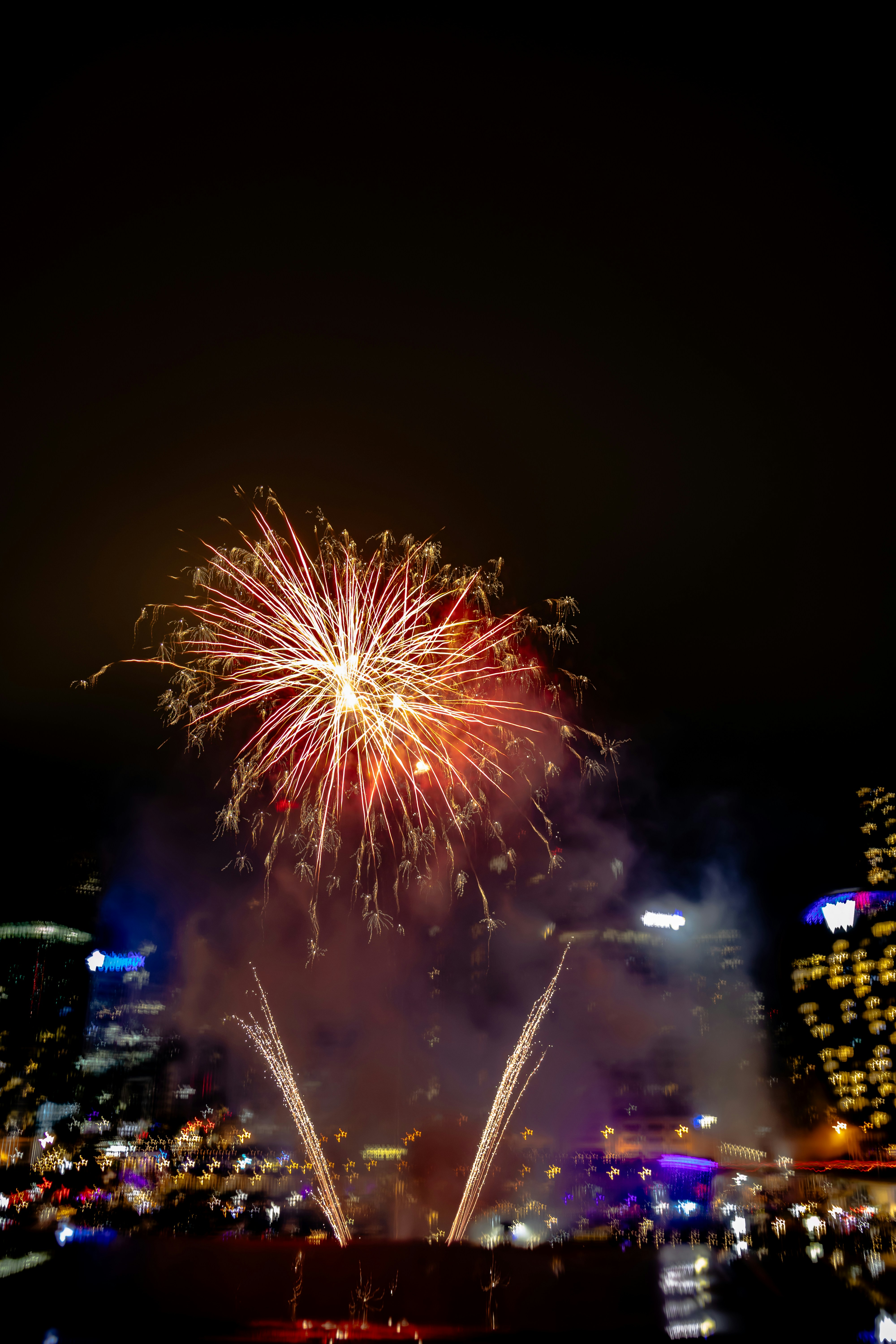 Fireworks explode in a vibrant display over a cityscape at night, with bursts of red, yellow, and white illuminating the sky. The colorful lights and reflections create a festive atmosphere, while city buildings and lights in the background add to the celebratory scene.Dohyuk You