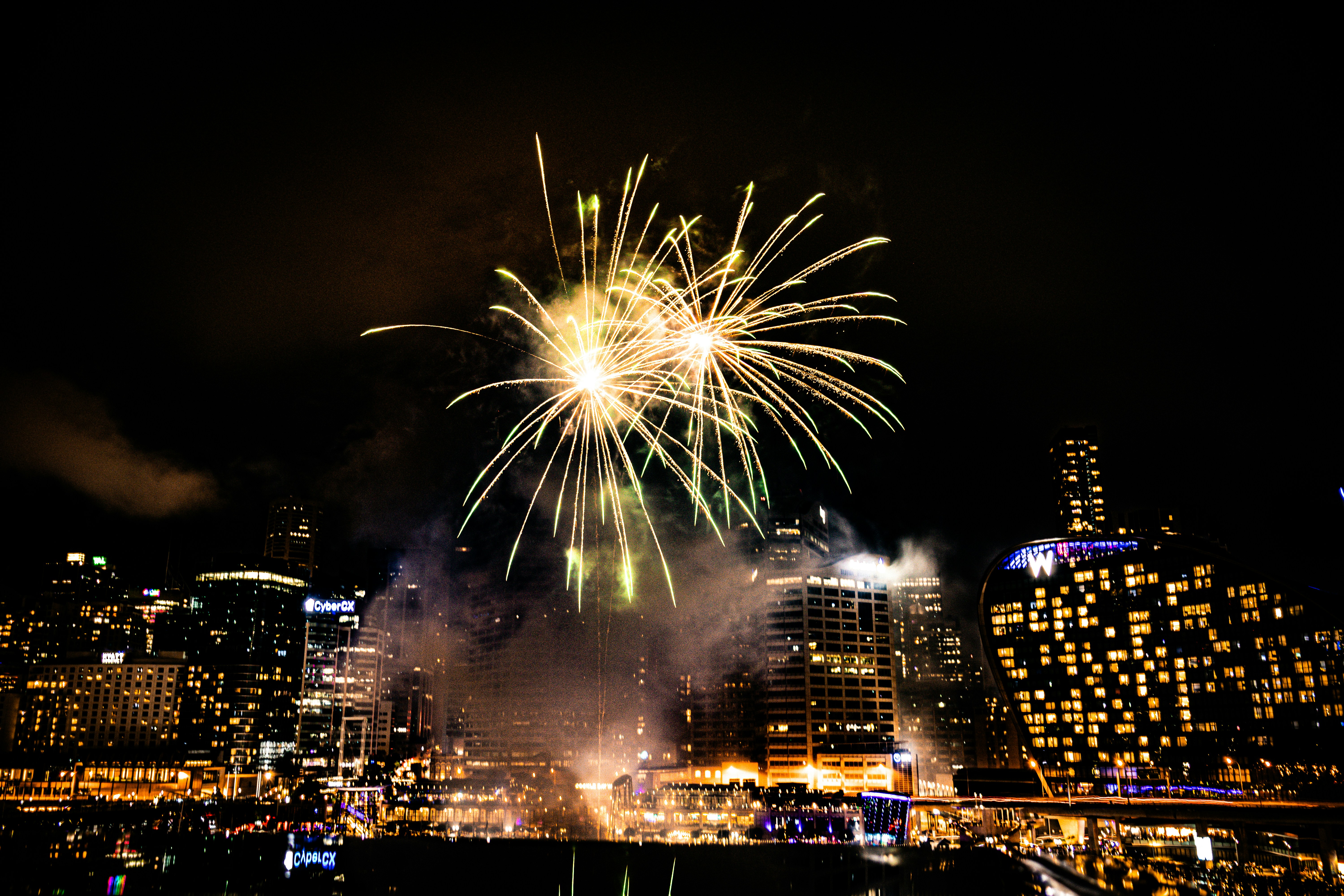 Fireworks burst in the night sky over a cityscape, illuminating the buildings and reflecting in the water below. The display features vibrant colors, primarily green and yellow, creating a festive and celebratory atmosphere. City lights and skyscrapers add to the lively scene, highlighting the urban celebration.Dohyuk You