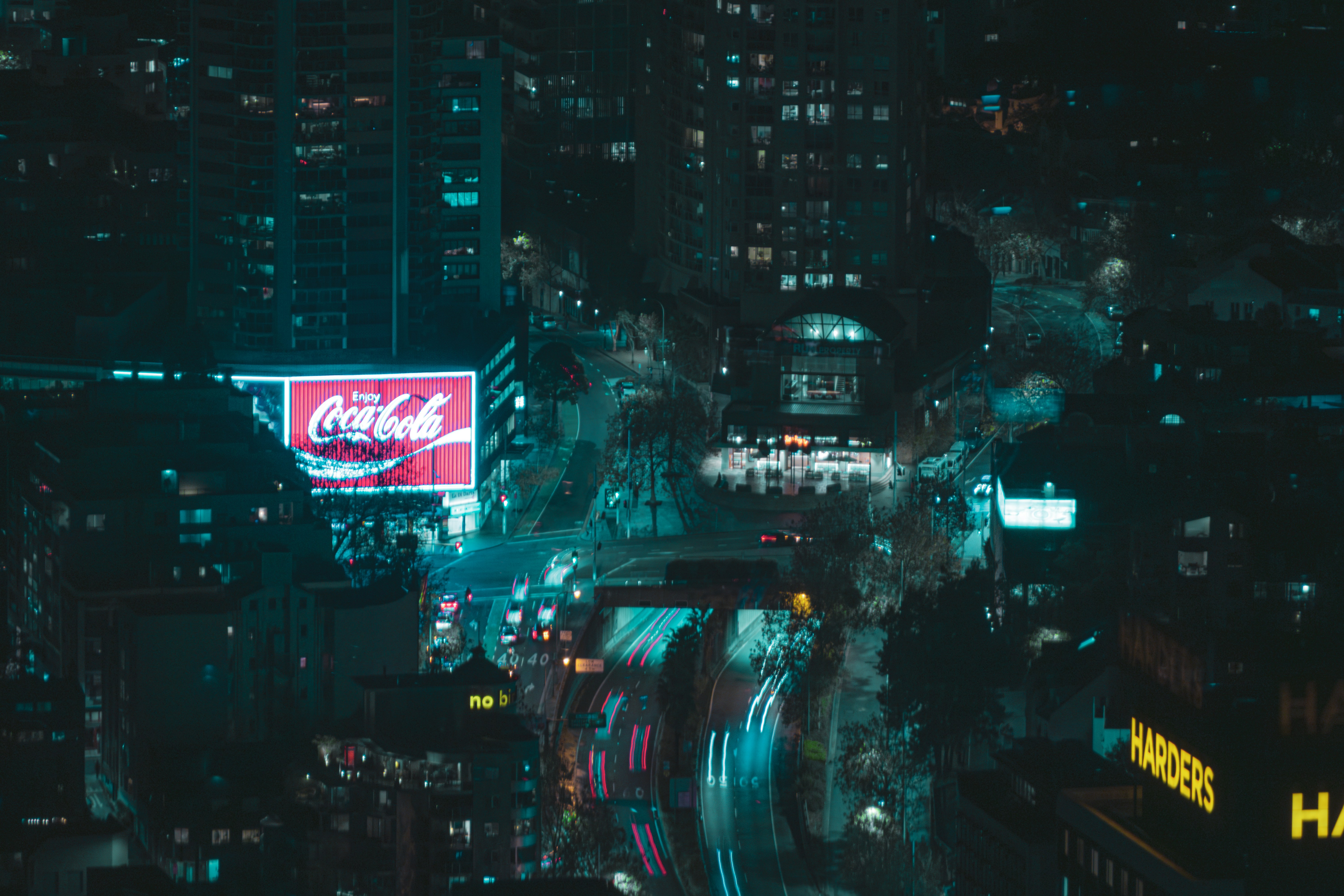 A bustling city street at night is illuminated by neon lights and the glow of a prominent Coca-Cola billboard. The long exposure captures the motion of cars, creating colorful light trails that add dynamism to the urban scene. High-rise buildings and trees frame the busy, vibrant nightlife of the city.