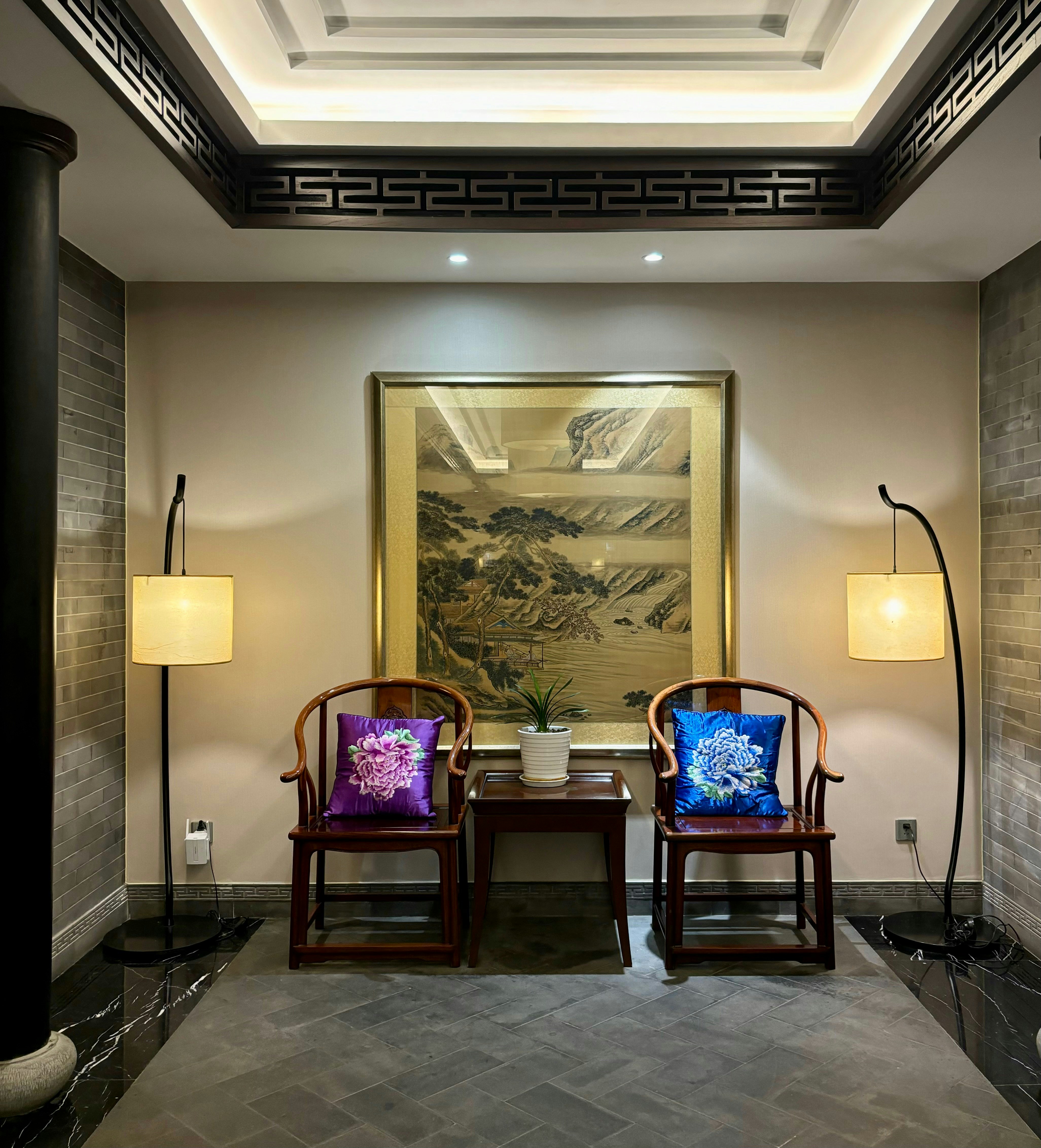 Two wooden chairs with floral cushions flank a small central table with a plant. A framed landscape painting dominates the wall, illuminated by warm lamps.