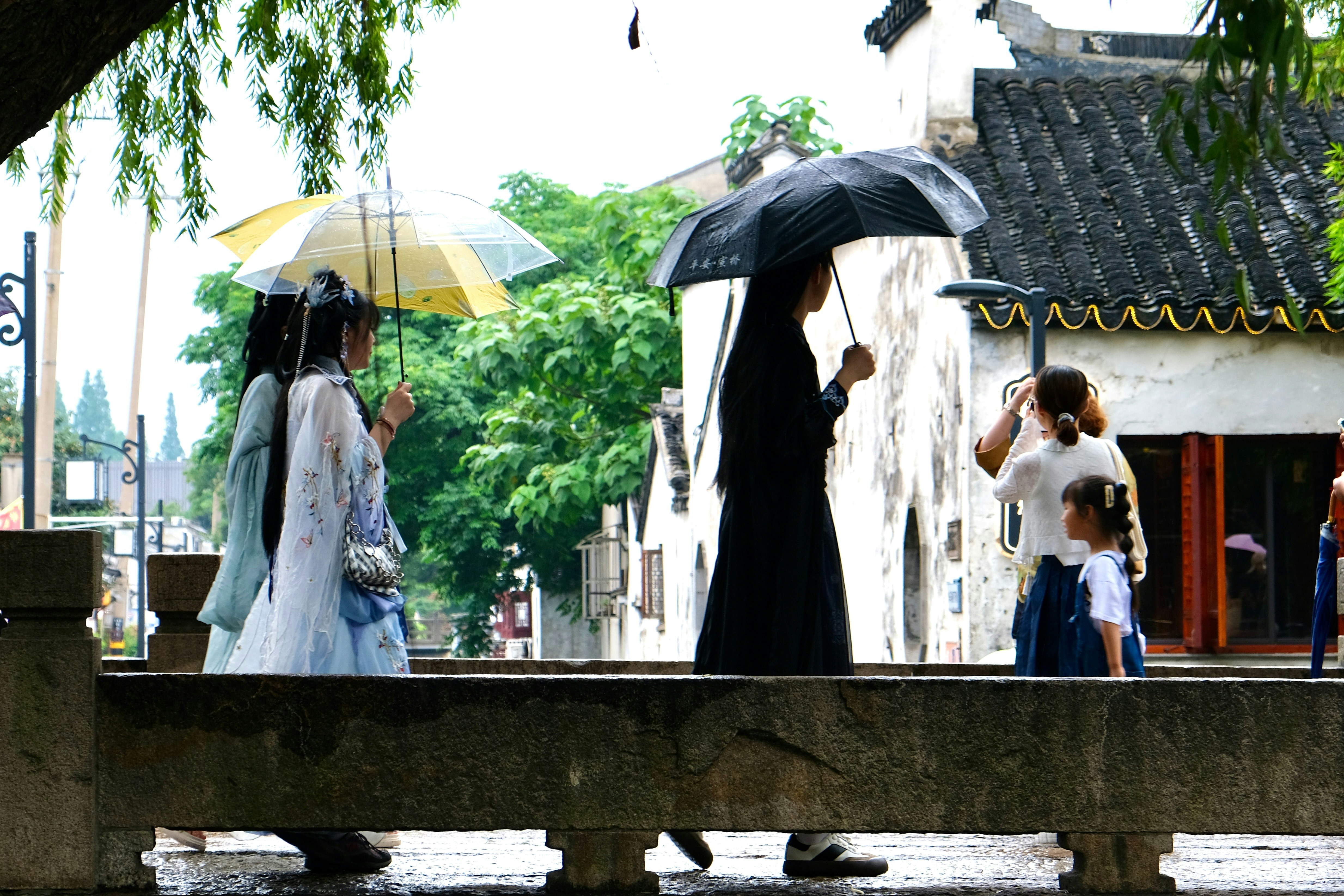 People with umbrellas crossing a stone bridge in a traditional setting on a rainy day.