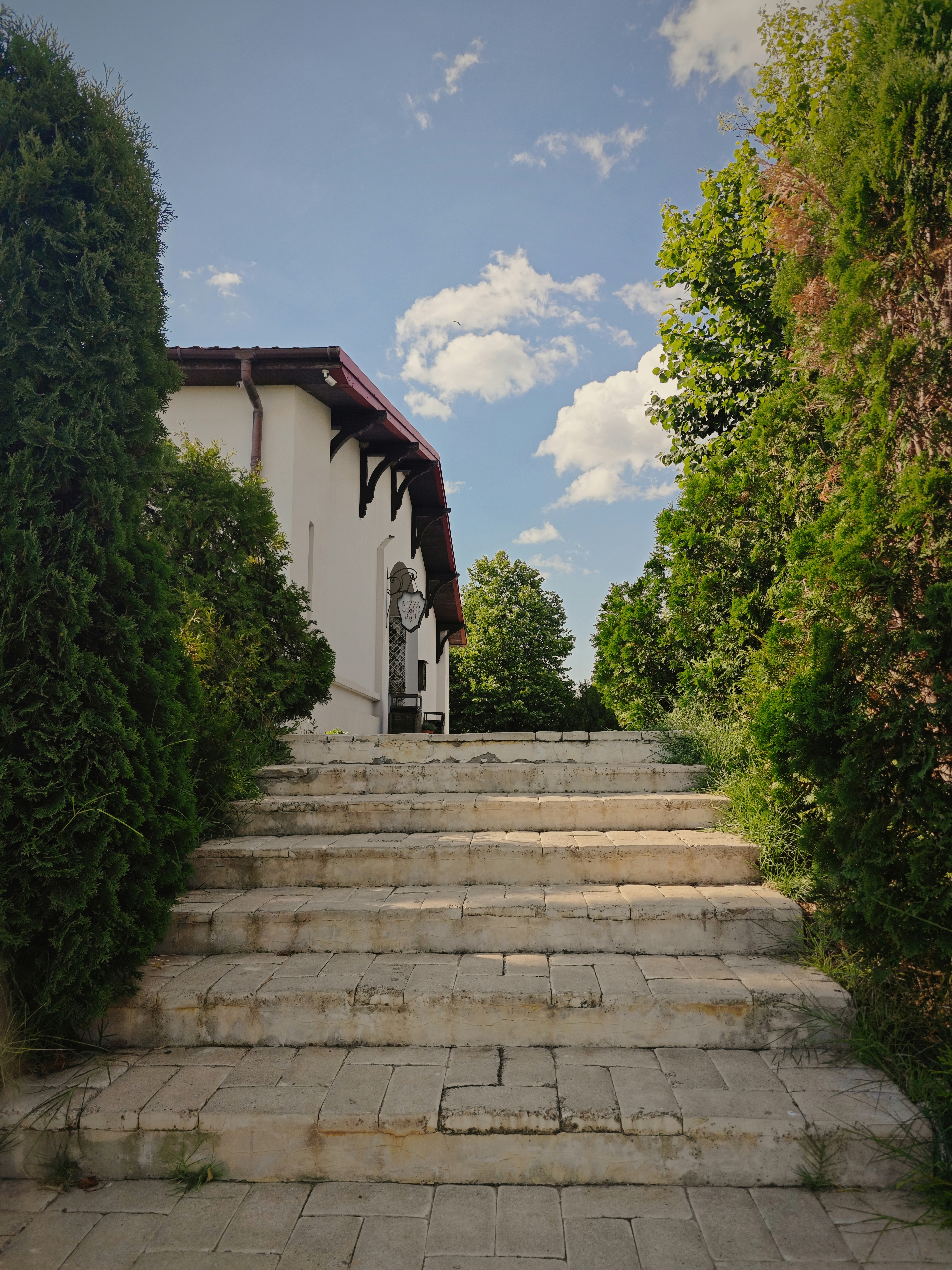 Stone steps rise toward a white building framed by evergreen hedges under a bright blue sky.