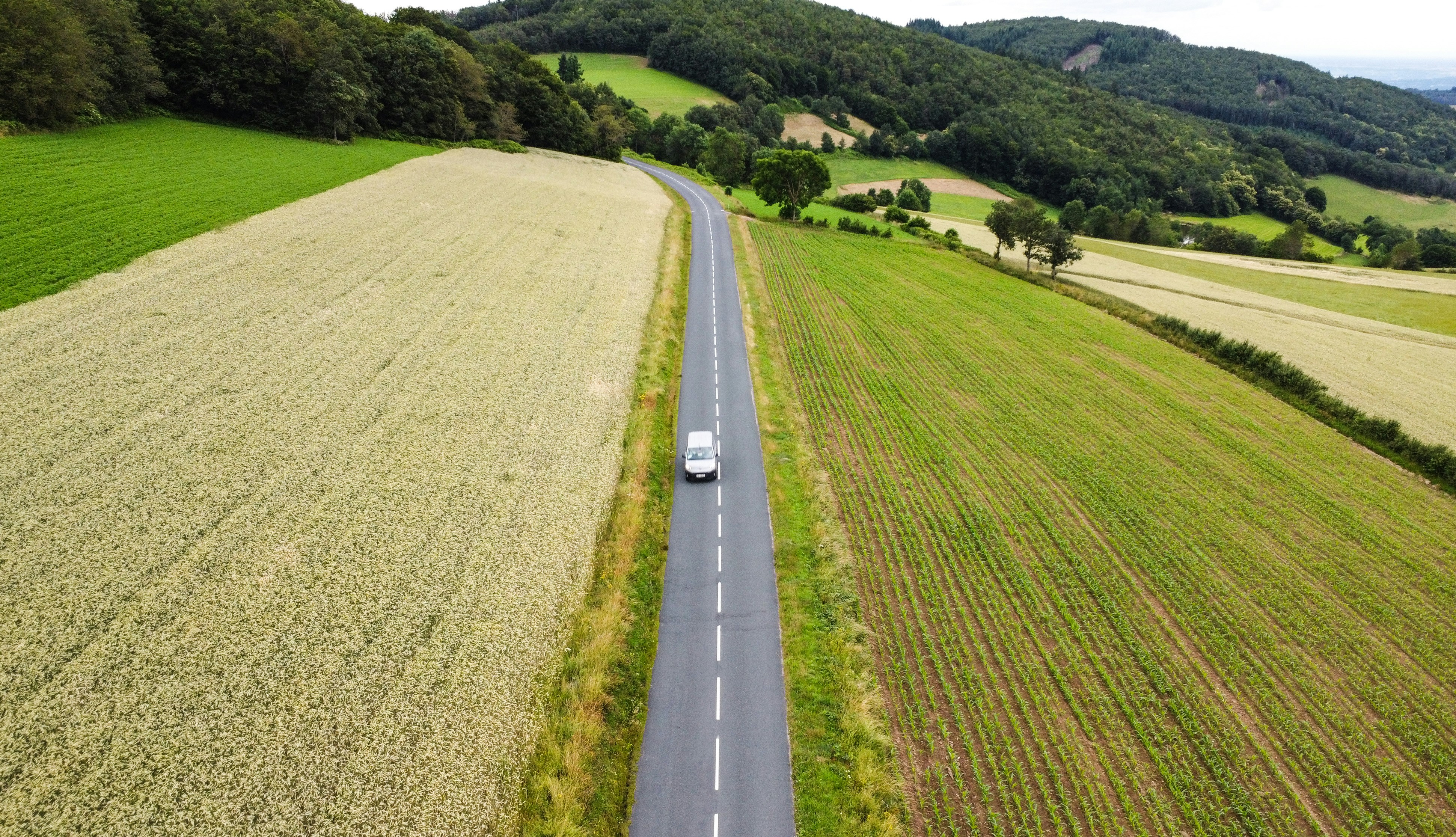Aerial photograph of a two-lane road slicing through patchwork fields, with a lone white car centered on the road.