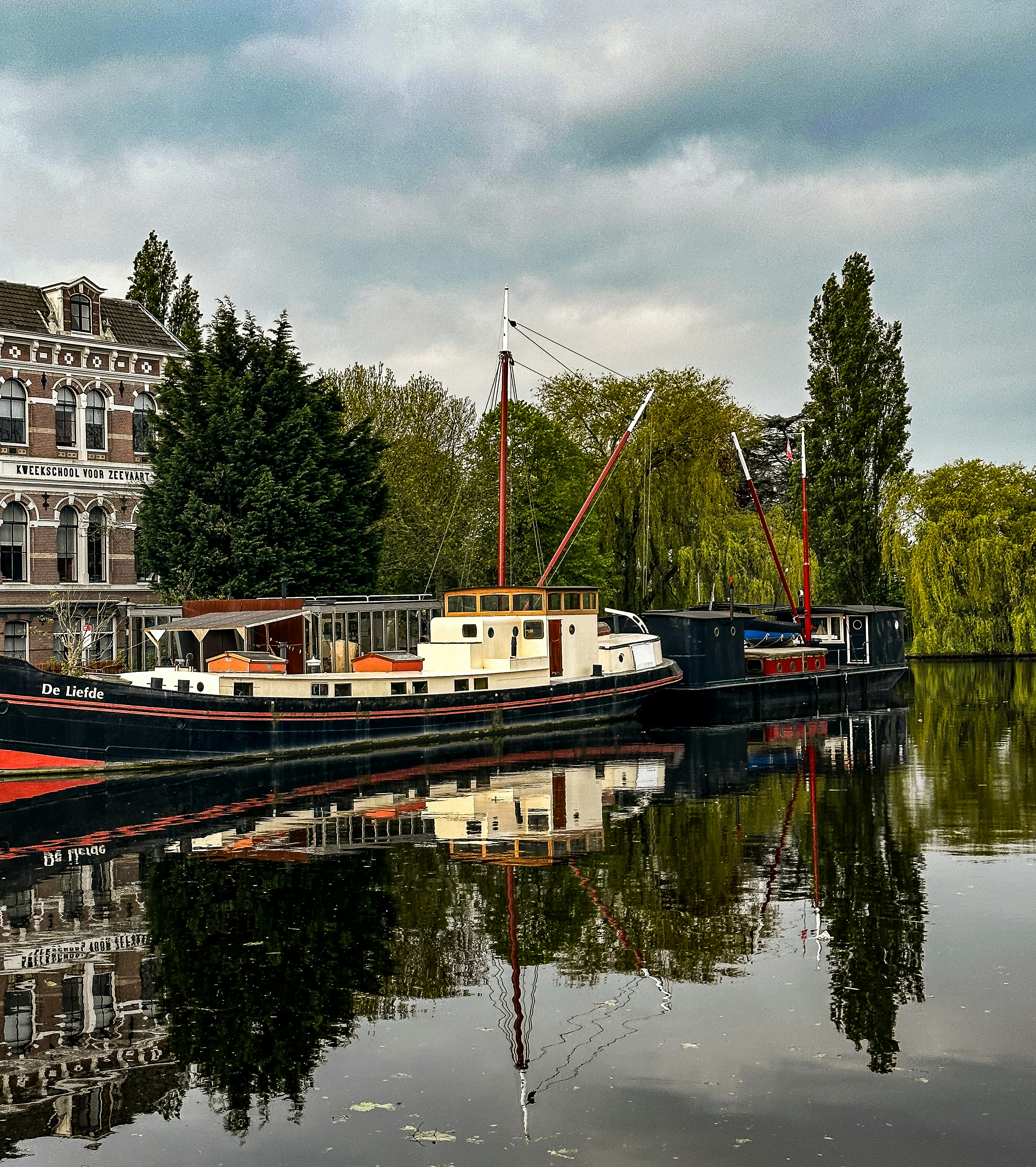 Historic boats moored along a calm canal with lush greenery and a classic building in the background.