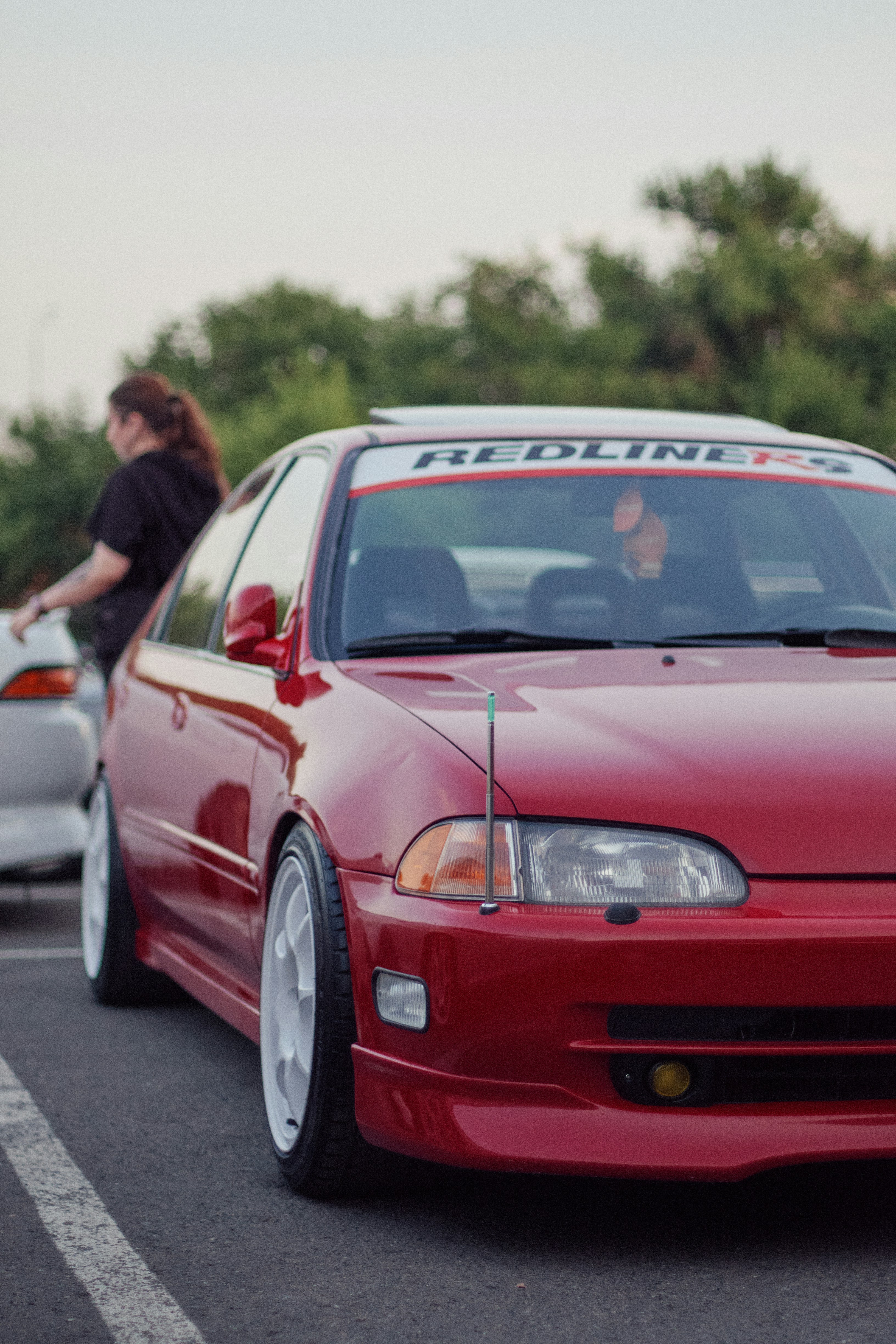 Redline Focus: A lowered red coupe with a slim green antenna at a daytime car meet