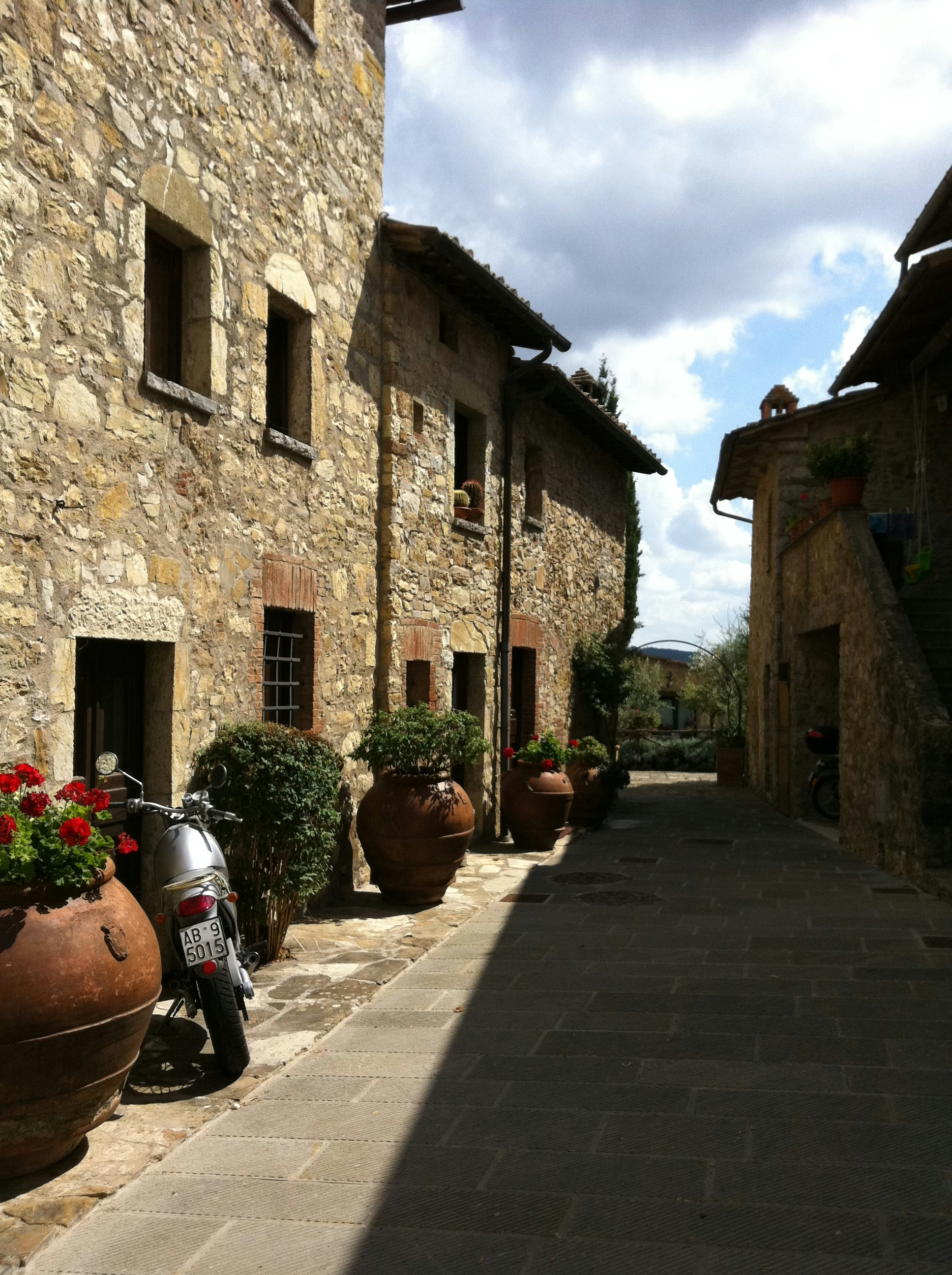 Sunlit stone alley lined with large terracotta pots and a parked motorcycle, extending toward a distant village. Warm textures and Mediterranean charm define the scene.