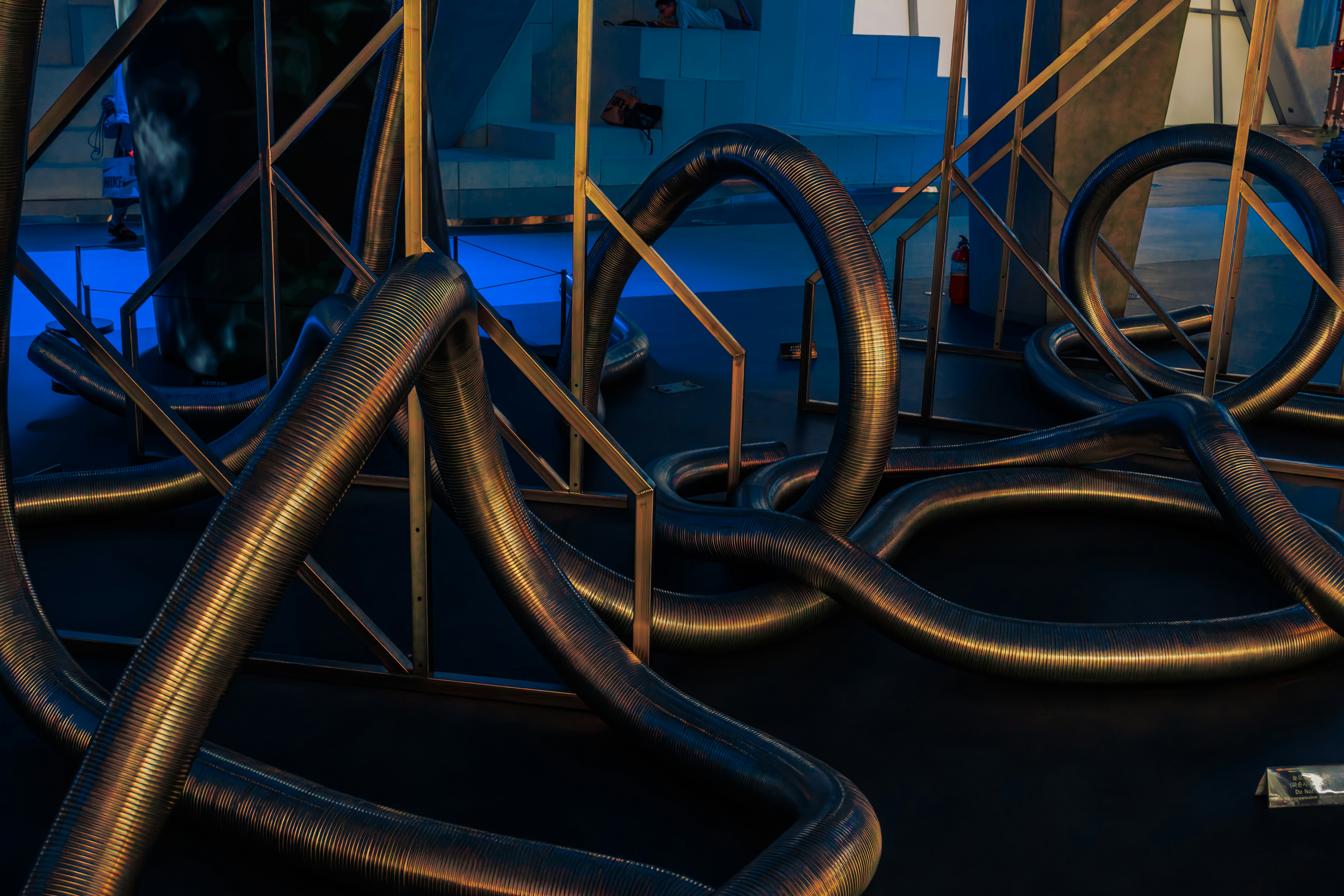 Metallic tubes intertwined in geometric frames under blue lighting, creating a futuristic atmosphere with dynamic reflections.