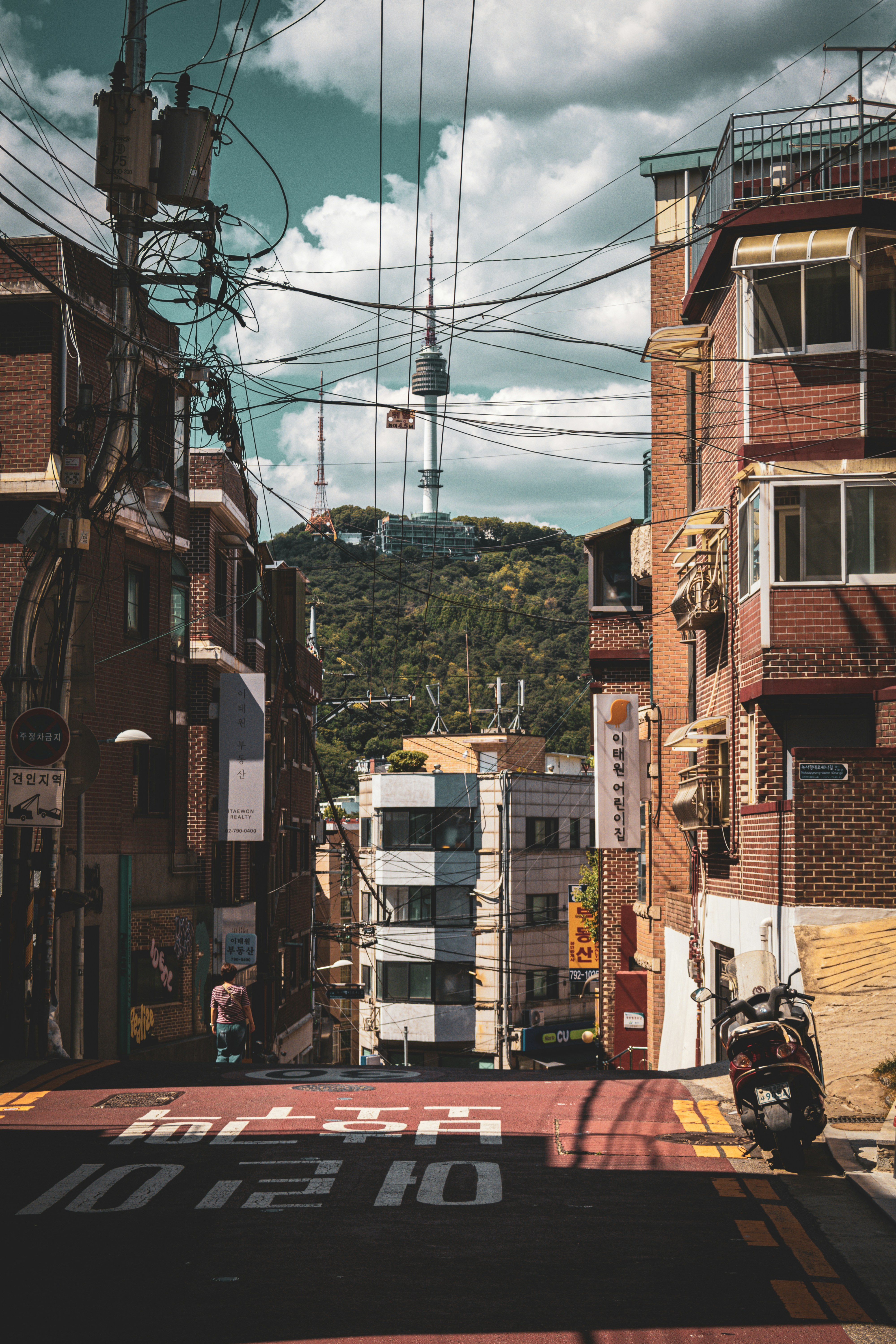 A steep street in Seoul with red brick buildings on either side, leading up to the iconic Namsan Seoul Tower. The road is marked with Korean text, and power lines create a web above. A scooter is parked on the right, emphasizing the urban vibe.Dohyuk You