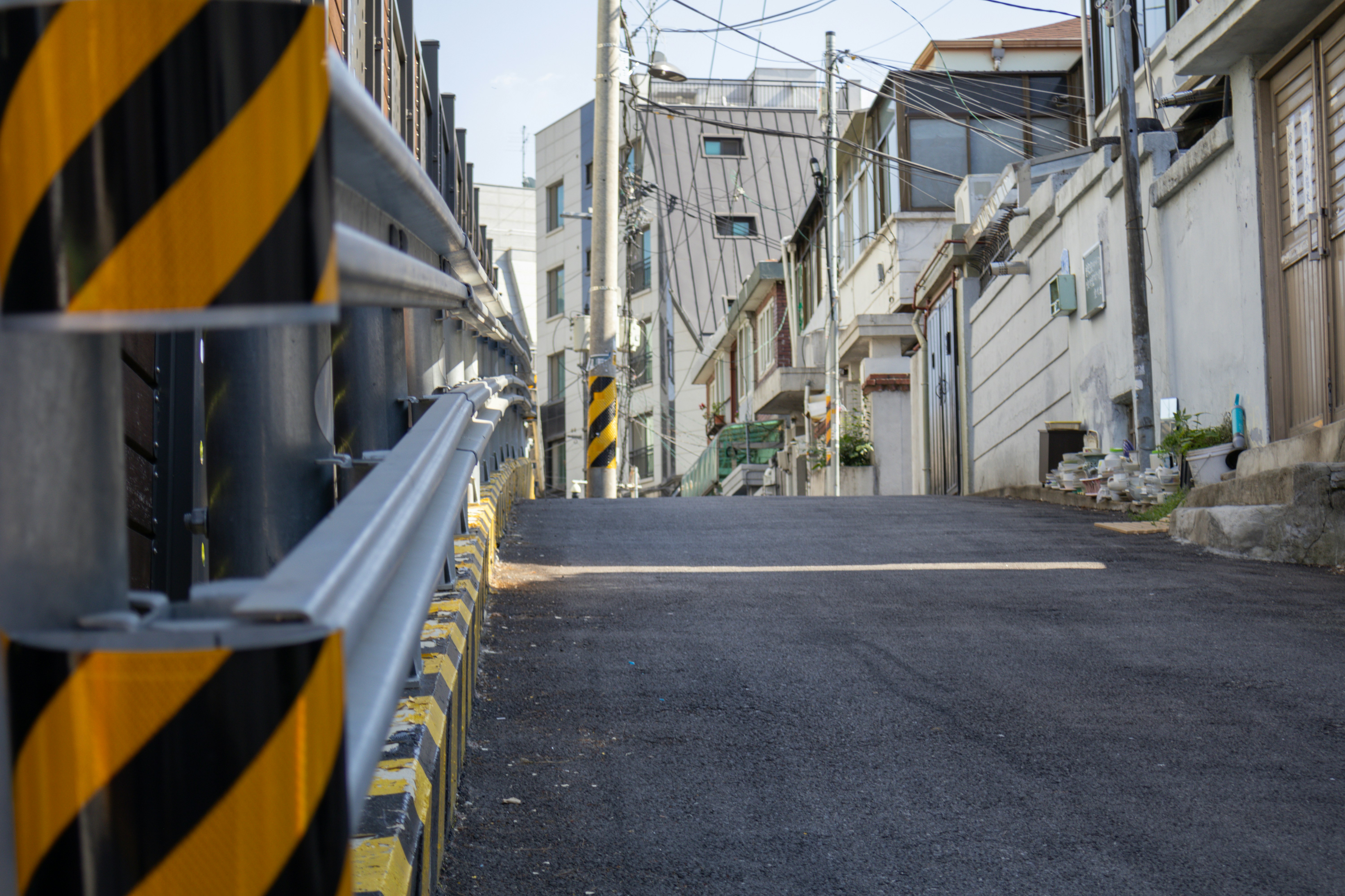 A steep, narrow street in a residential area with safety barriers featuring yellow and black stripes. The houses are closely packed, and utility poles with overhead wires add to the urban atmosphere. The clear blue sky and sunlight highlight the clean pavement.Dohyuk You
