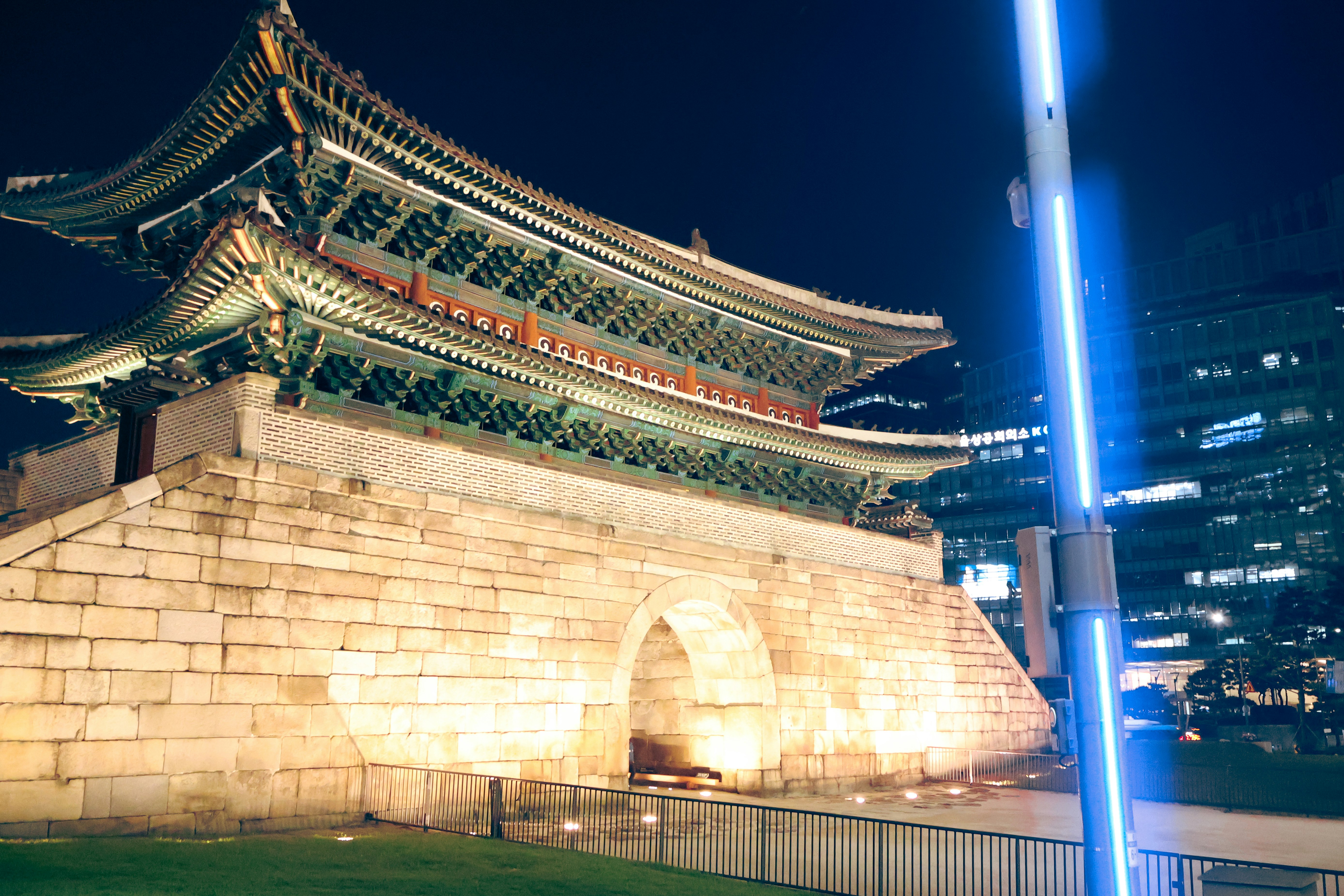 A traditional Korean gate, illuminated at night, stands in stark contrast to the modern buildings surrounding it. The intricate architecture of the gate is highlighted by warm lighting, while a blue neon light adds a modern touch to the scene.