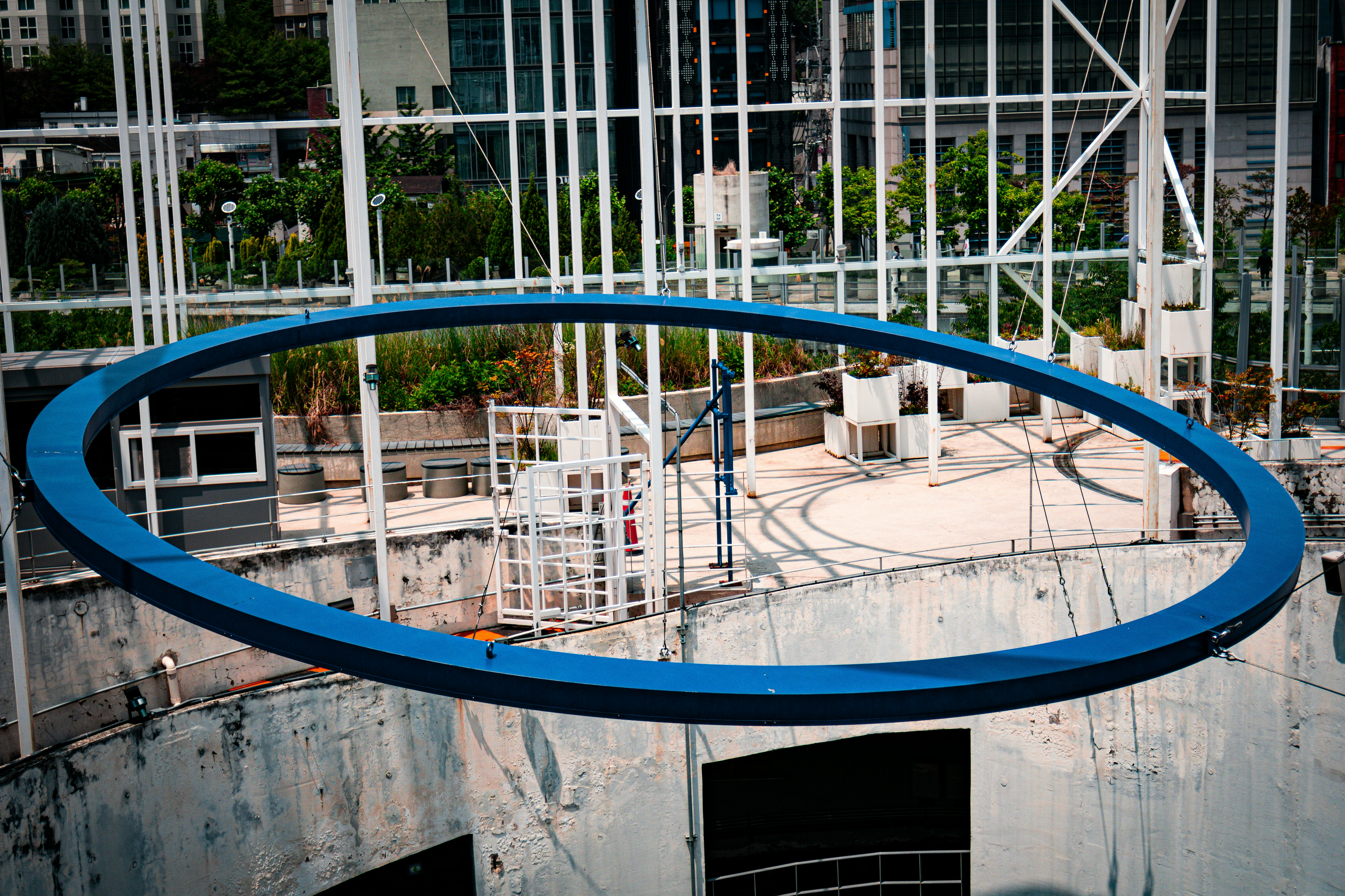 A rooftop installation featuring a large blue circular structure suspended above an urban garden. The area is surrounded by white metal beams and various potted plants, blending industrial and natural elements against a backdrop of city buildings.