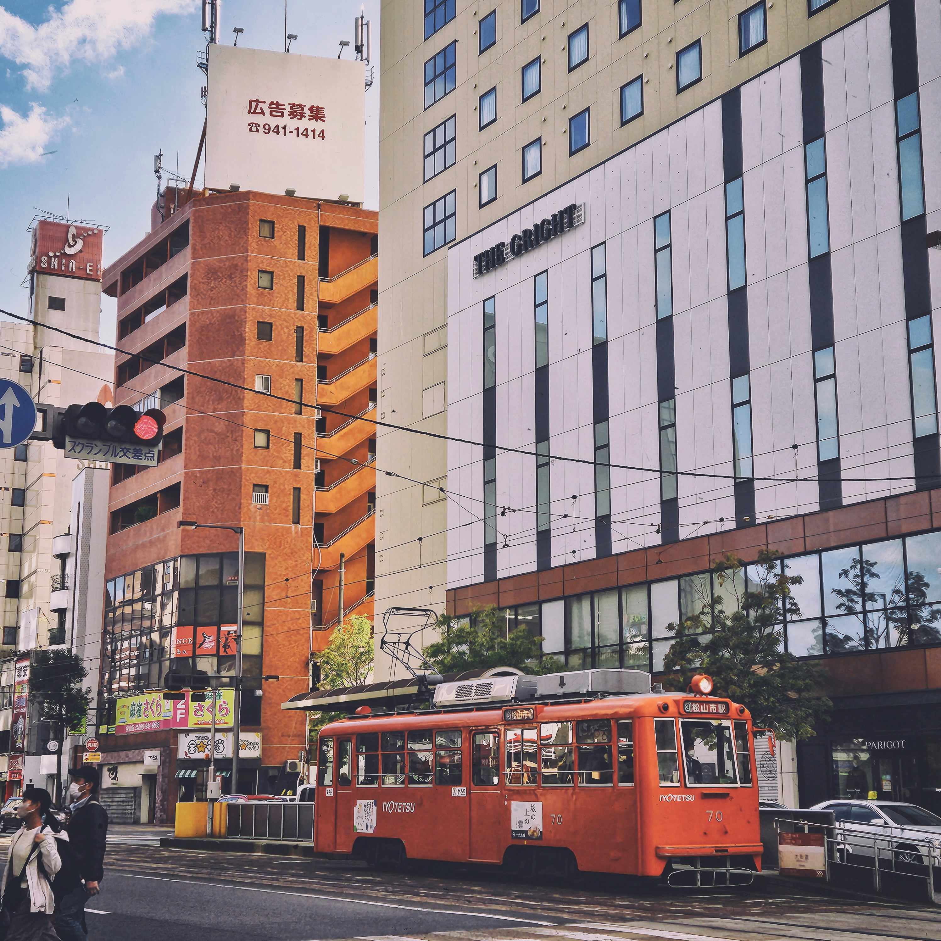 Photo capturing a red tram cutting through a sunlit urban corridor framed by glass towers and brick buildings.
