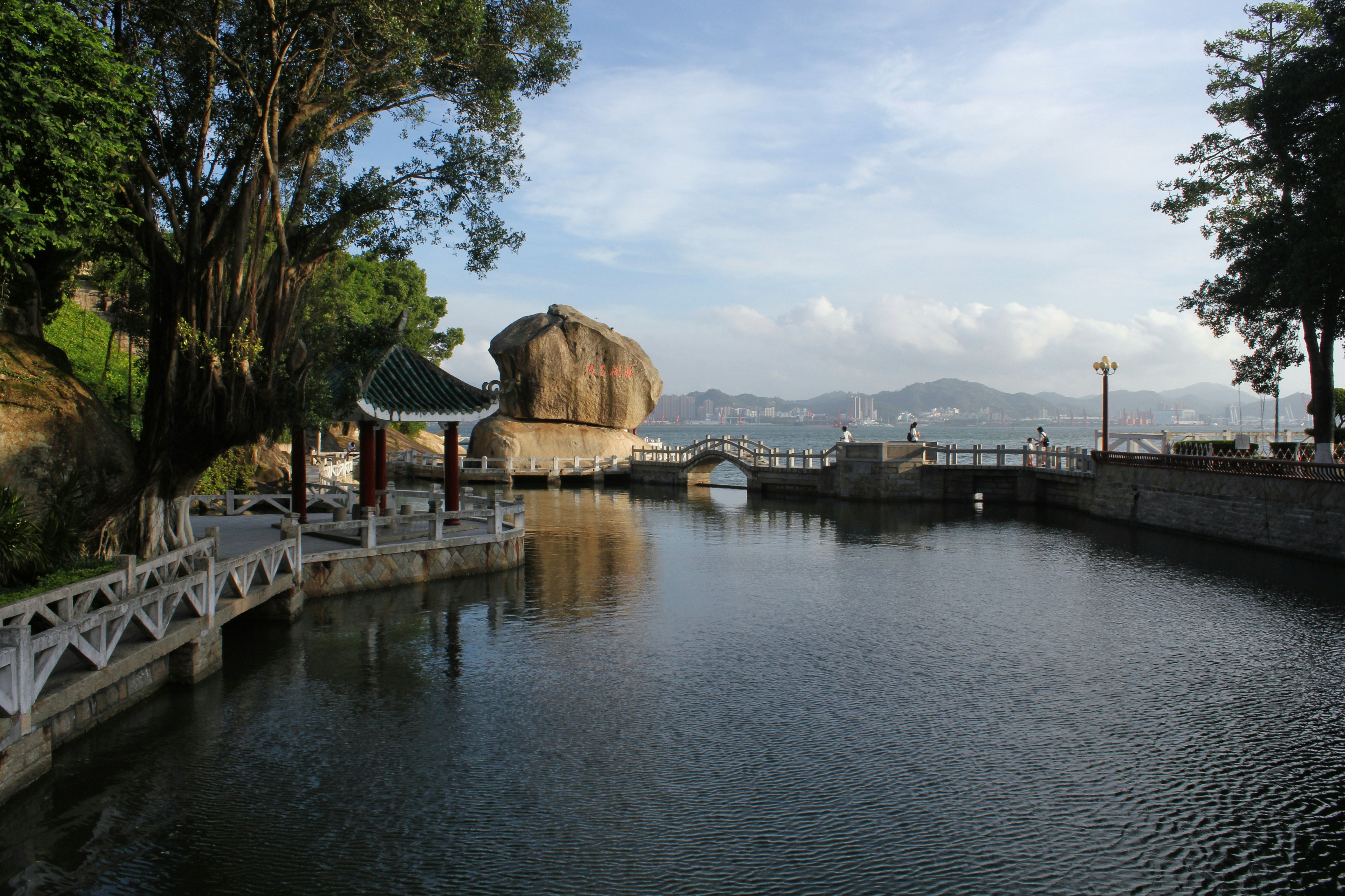 Stone Sentinel Over Calm Waters: Riverside Rock, Bridge, and Pavilion