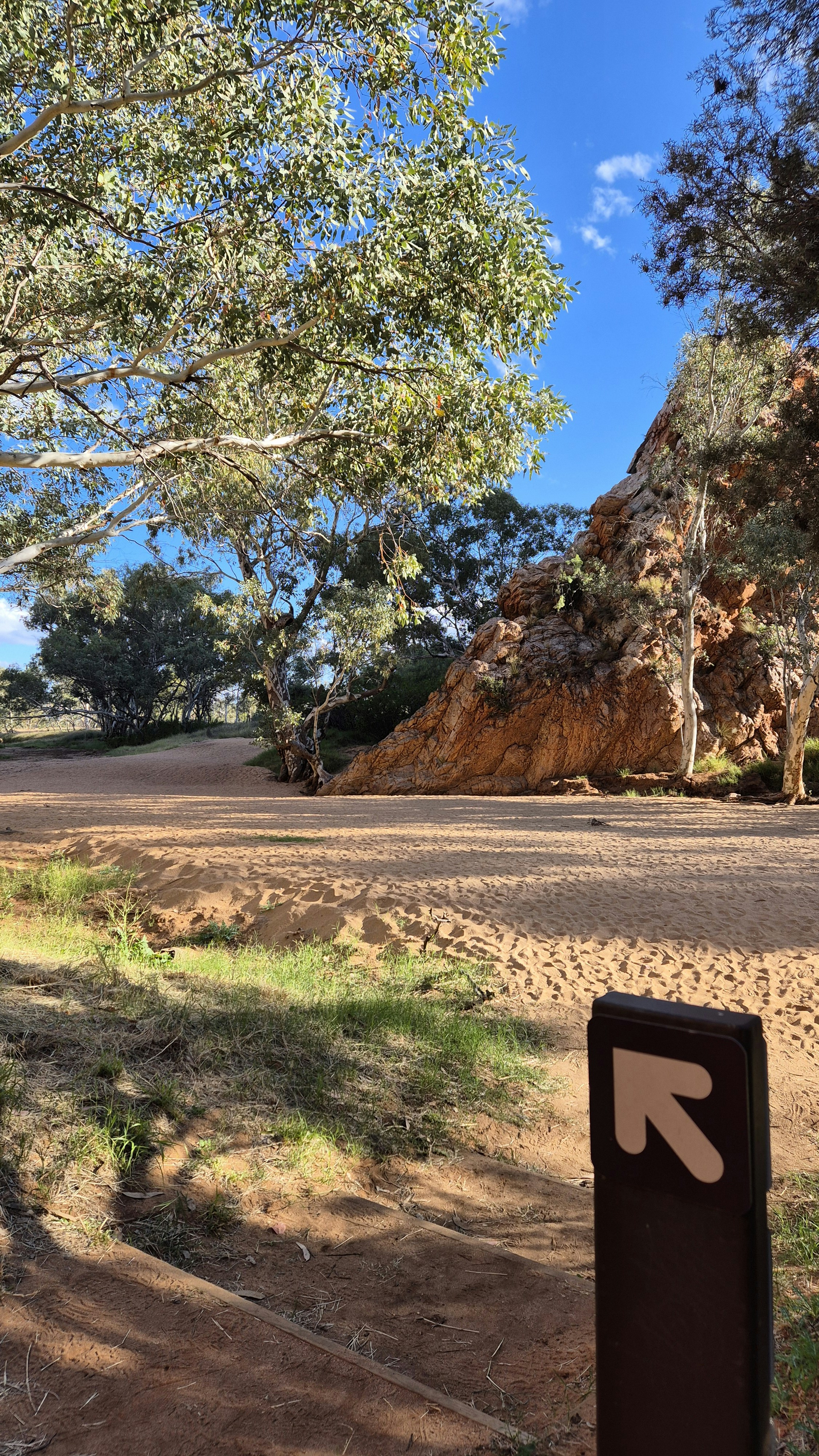 A directional sign points towards a sandy trail surrounded by lush greenery and rocky formations under a clear blue sky.