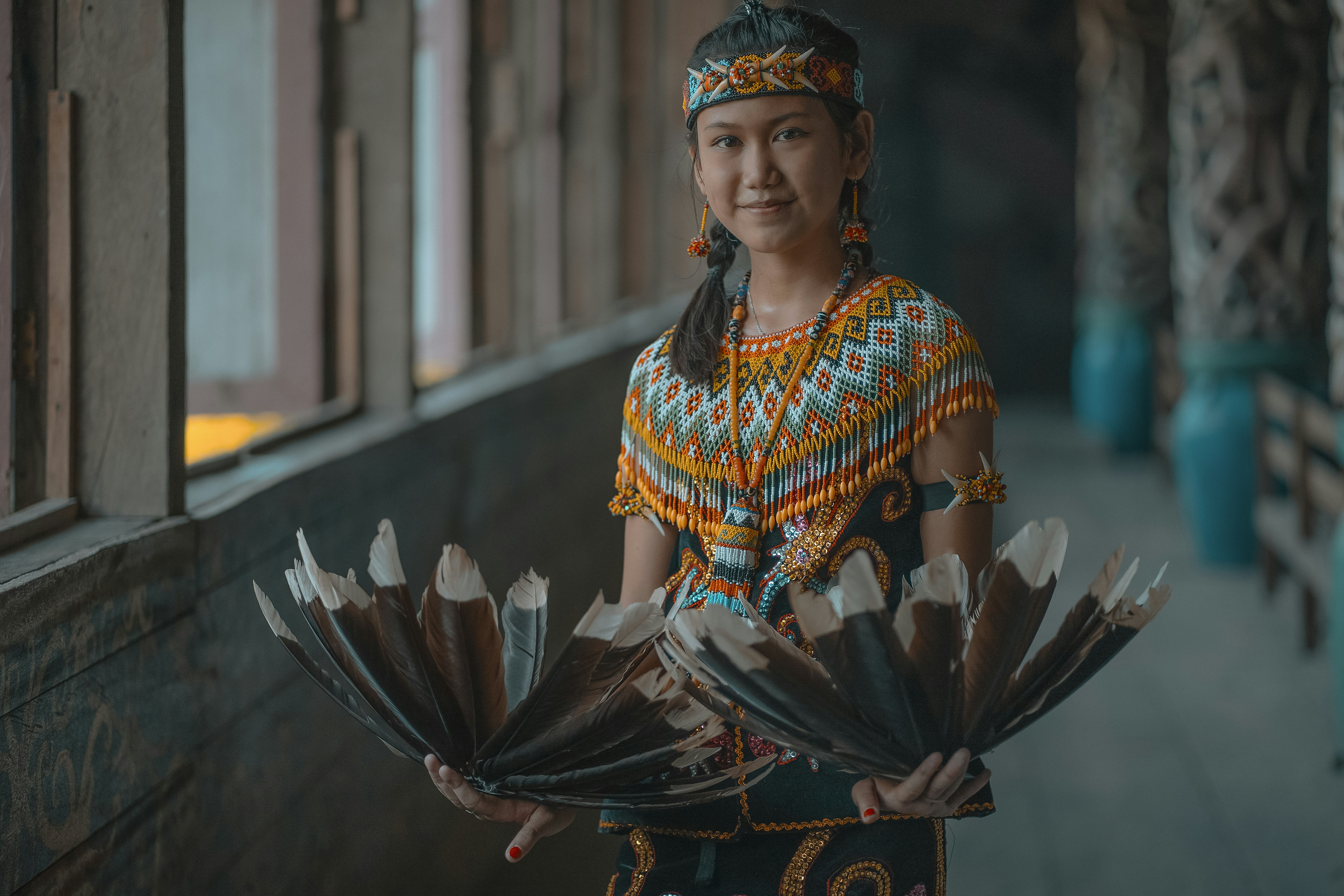 Young girl adorned in traditional attire holds decorative fans, showcasing her cultural heritage in a dimly lit corridor.