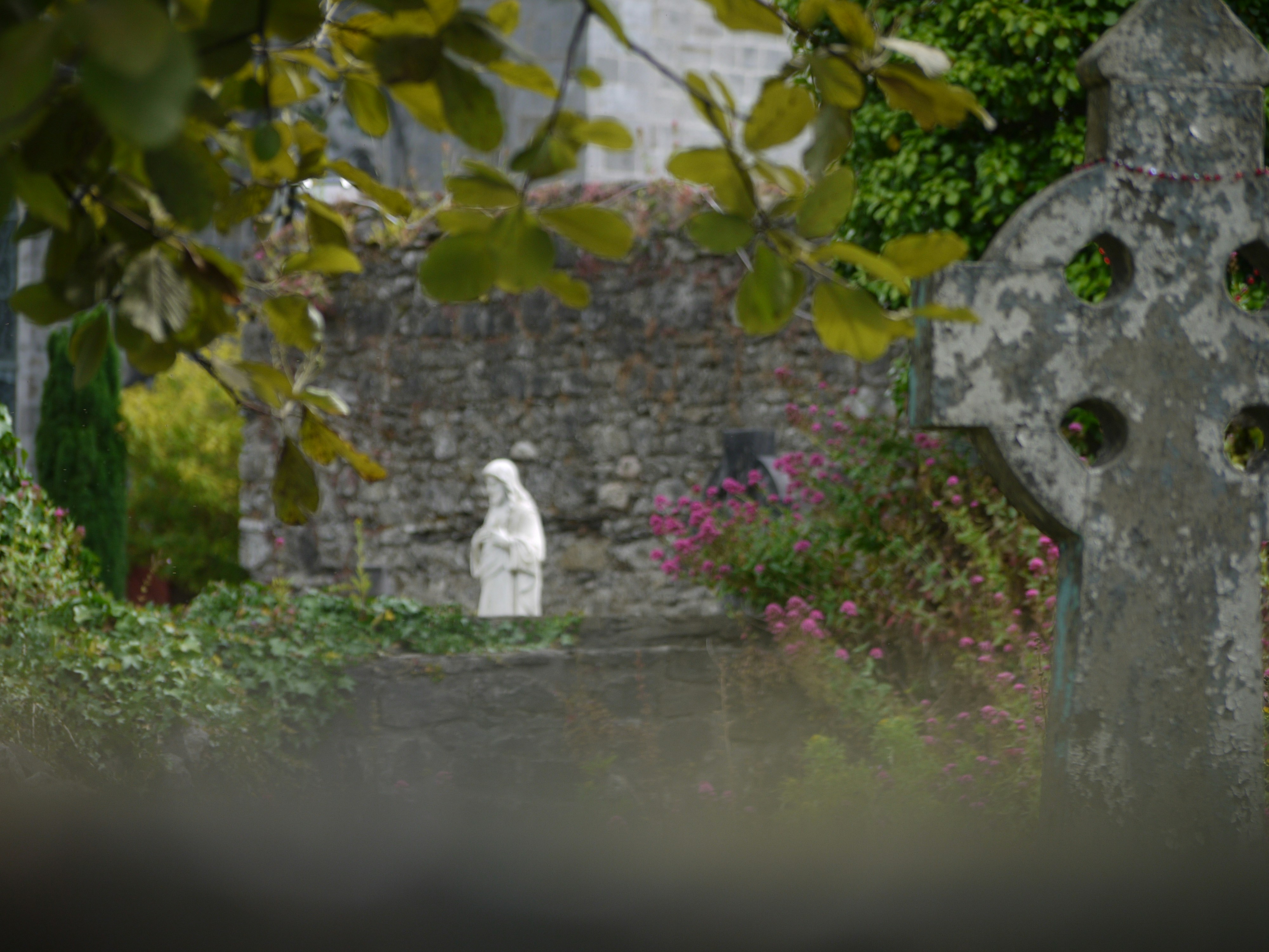 A white statue stands in a lush garden, framed by vibrant flowers and an ancient stone cross, evoking a sense of tranquility and history.