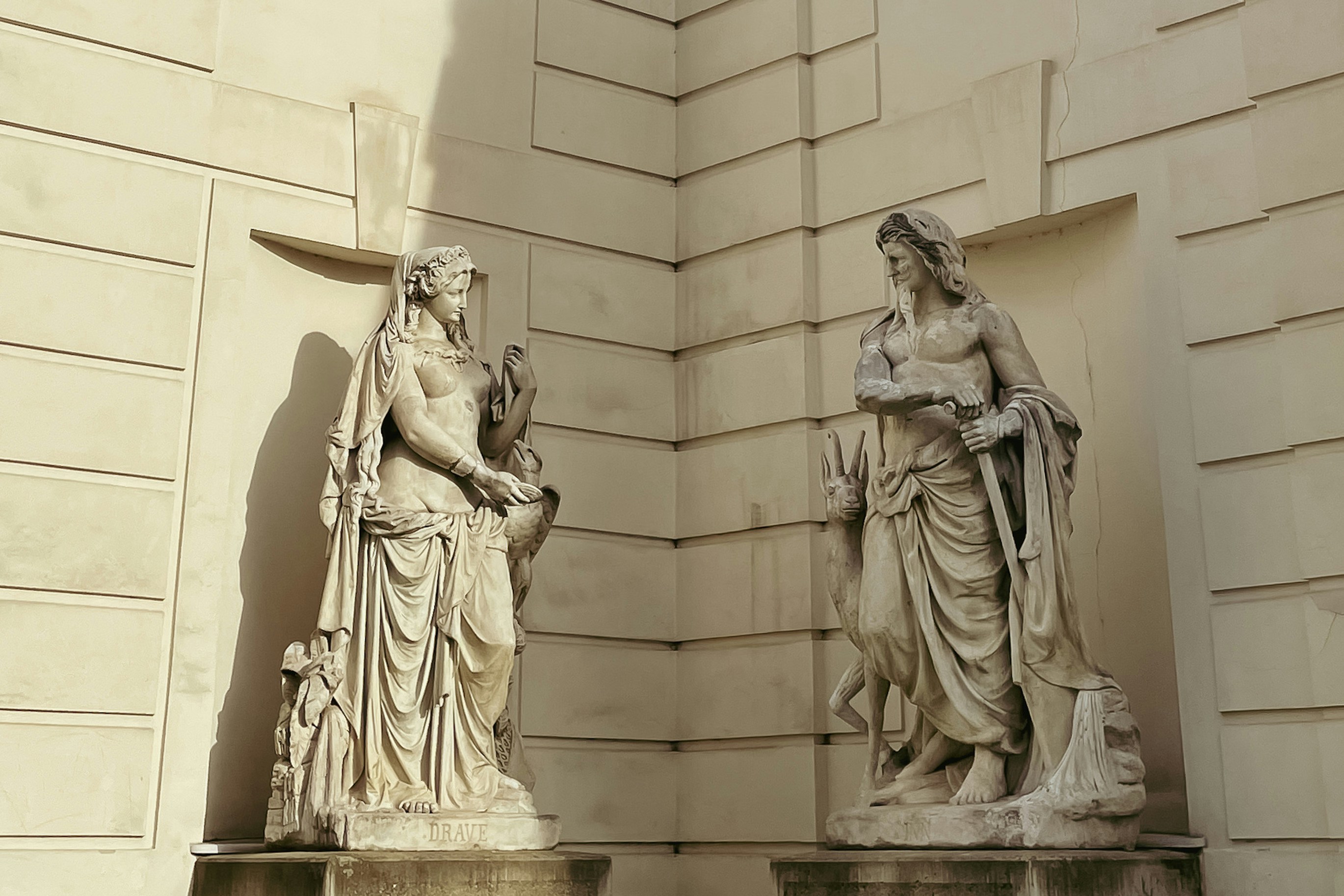 Two classical statues set against a geometric stone wall, illuminated by soft sunlight.