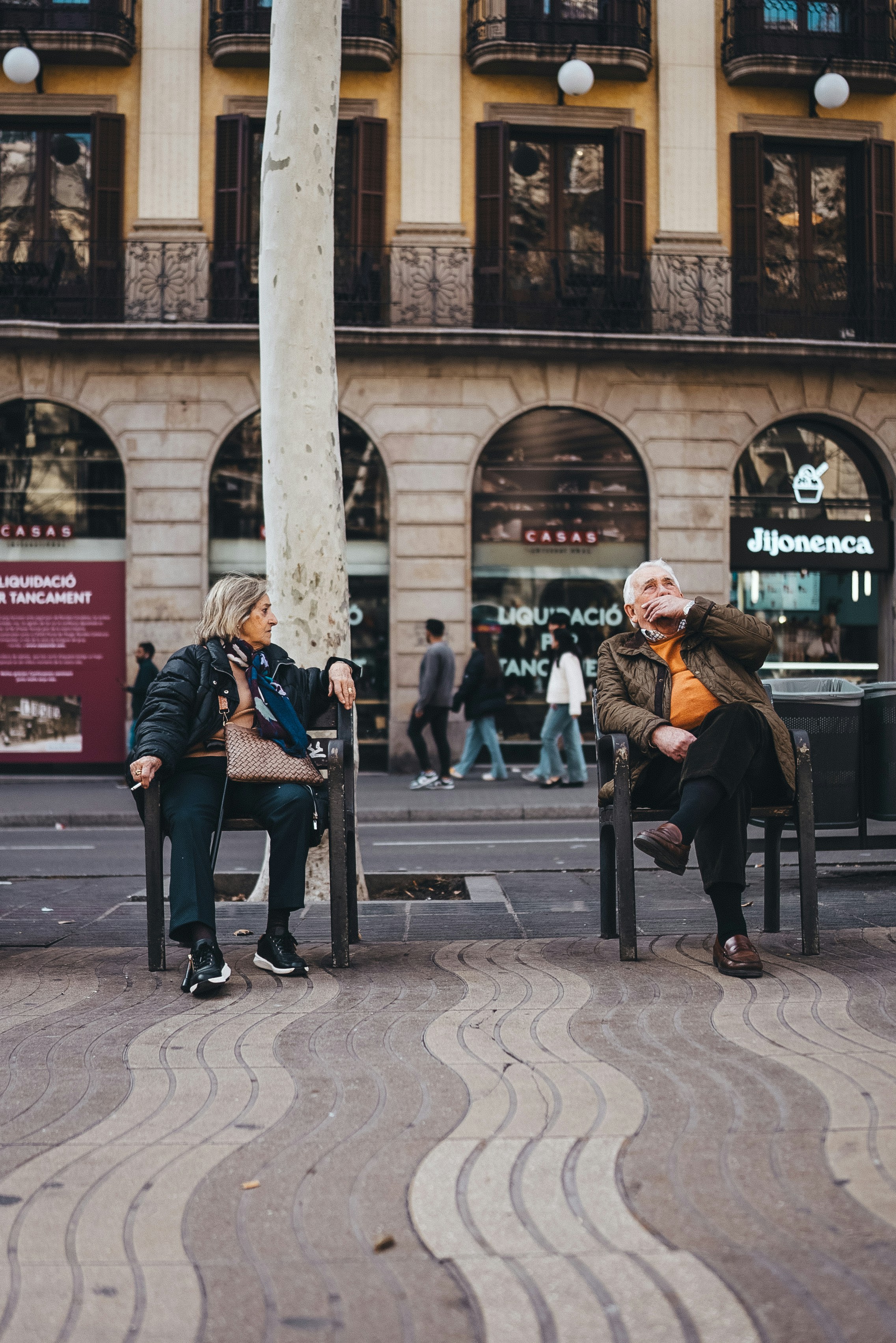 Two elderly pedestrians sit in vintage chairs along a tiled street beneath arched façades, with passersby moving in the background.