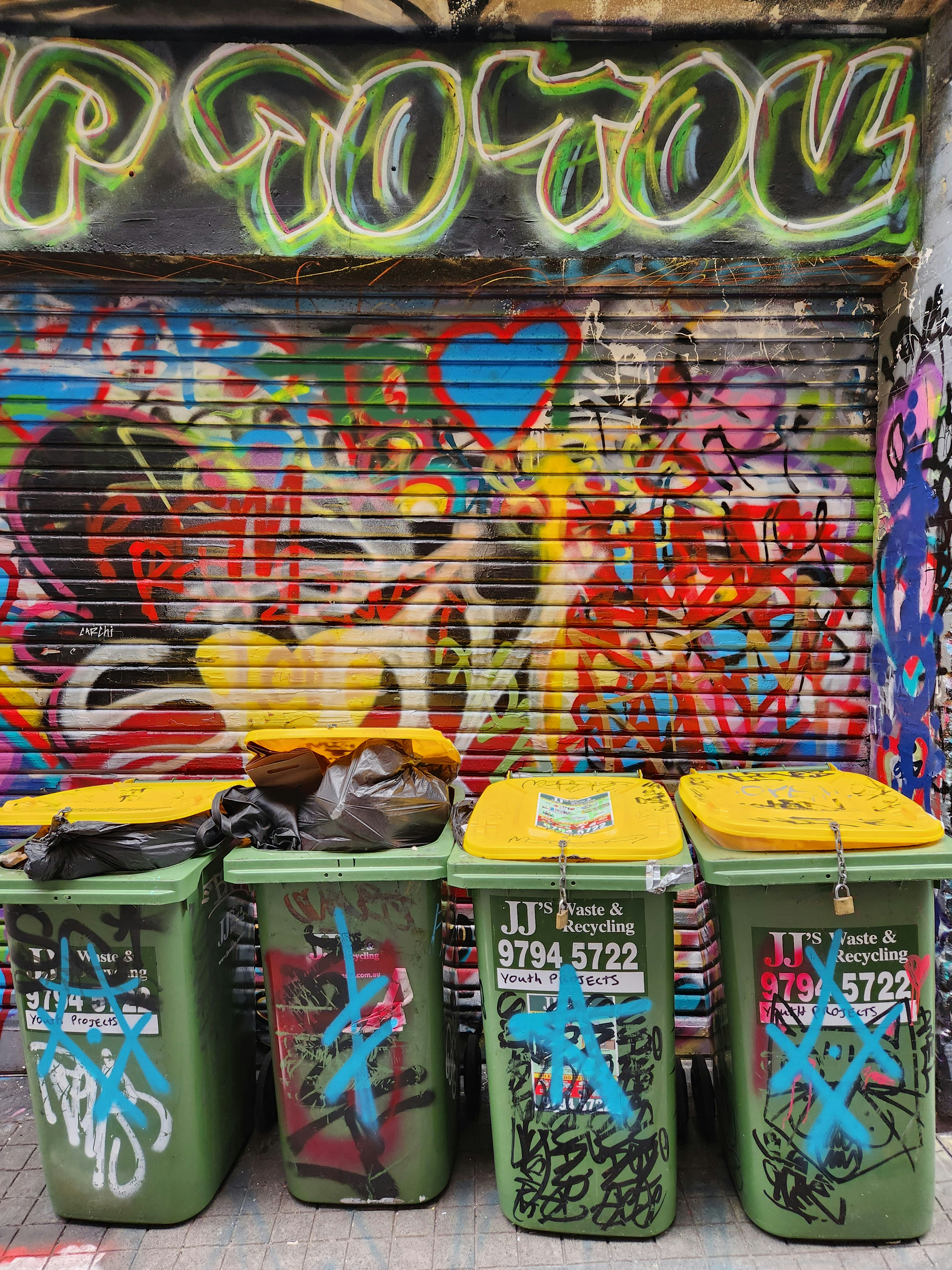 Colorful graffiti envelops a row of green recycling bins, showcasing a vibrant urban landscape. The interplay of art and waste highlights the city's creative spirit.