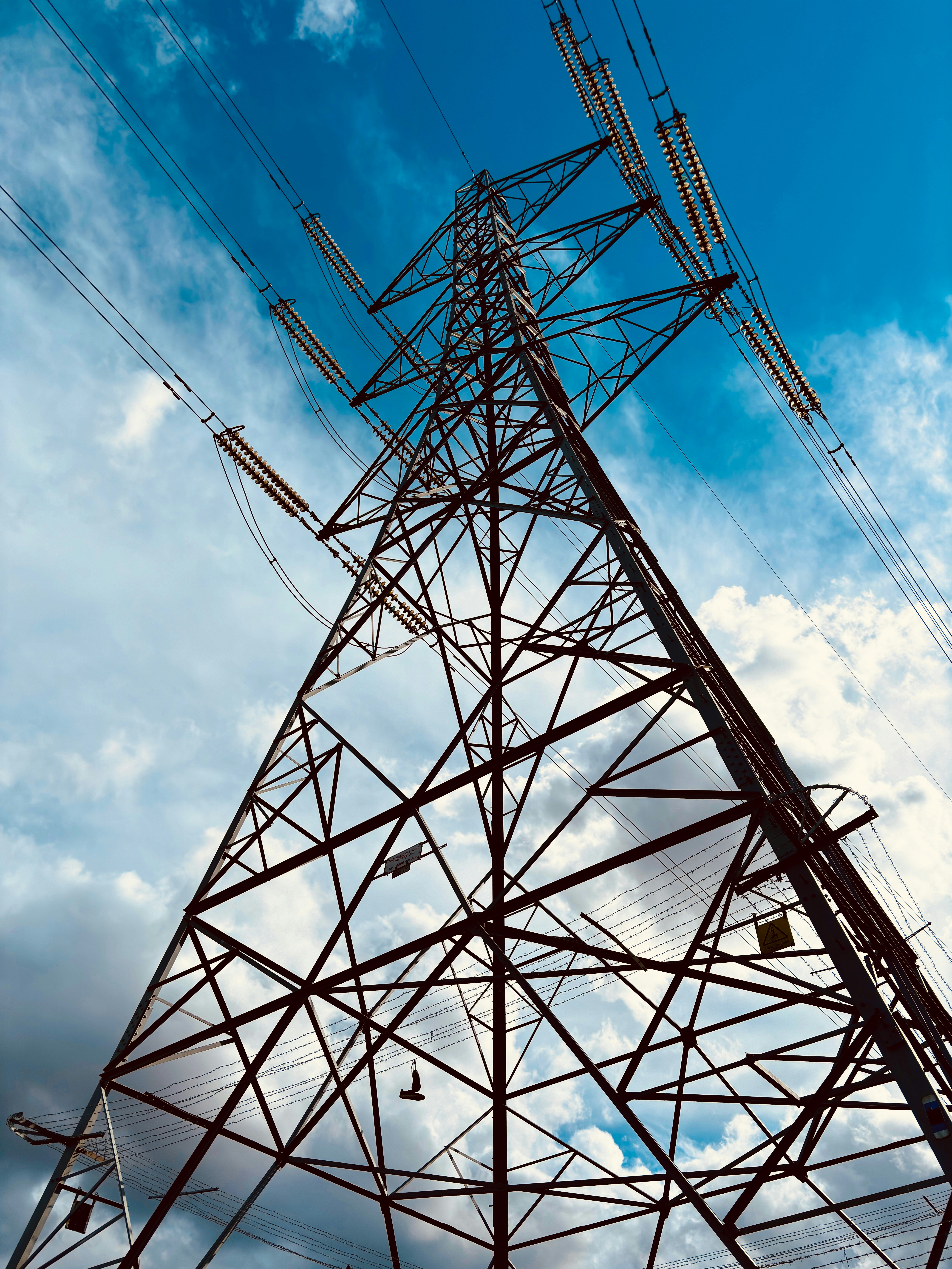 A high-voltage power tower stretches upward against a backdrop of dynamic clouds and blue sky.