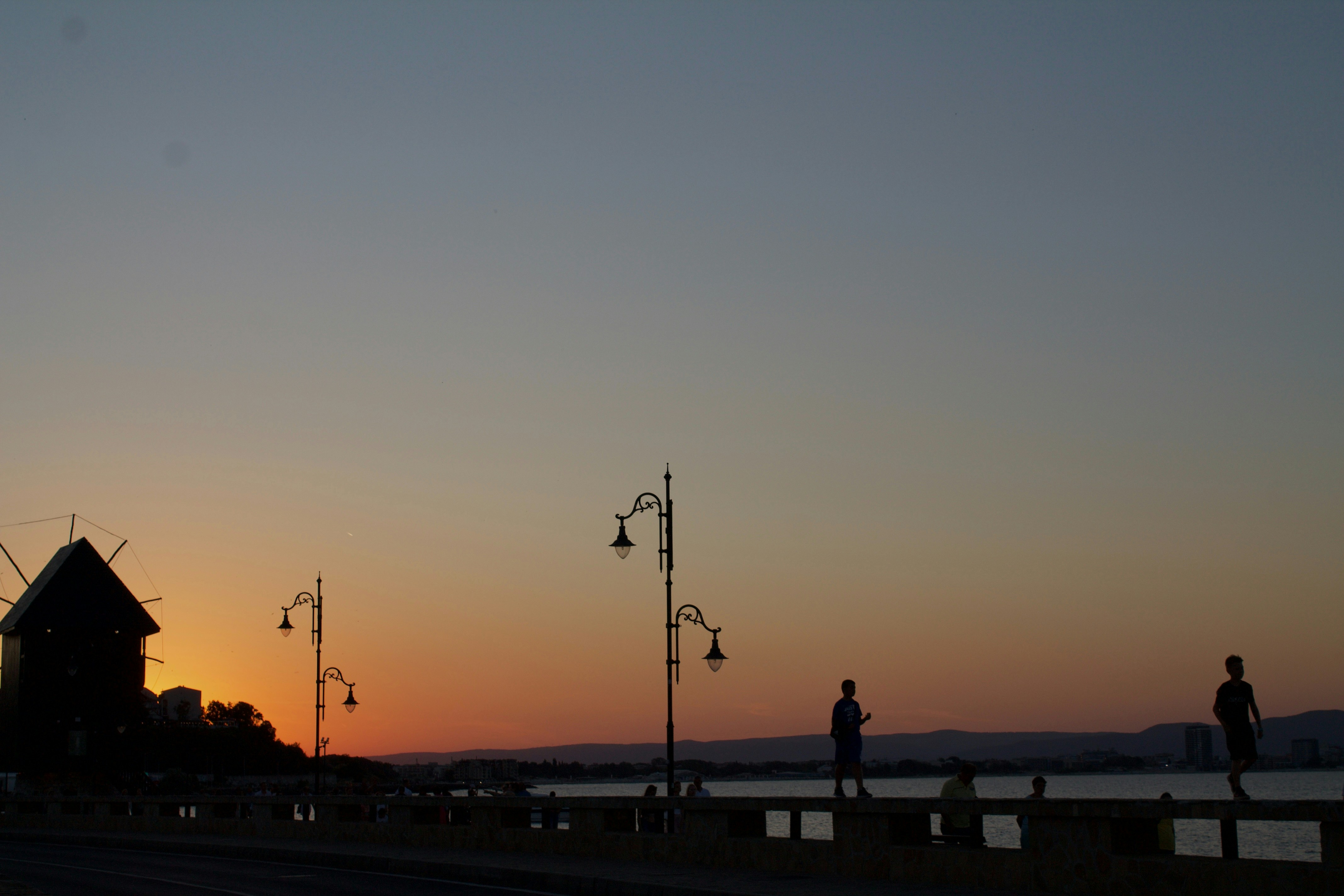 Silhouetted figures stroll along the waterfront as the sun sets behind a historic windmill, casting warm hues across the sky.