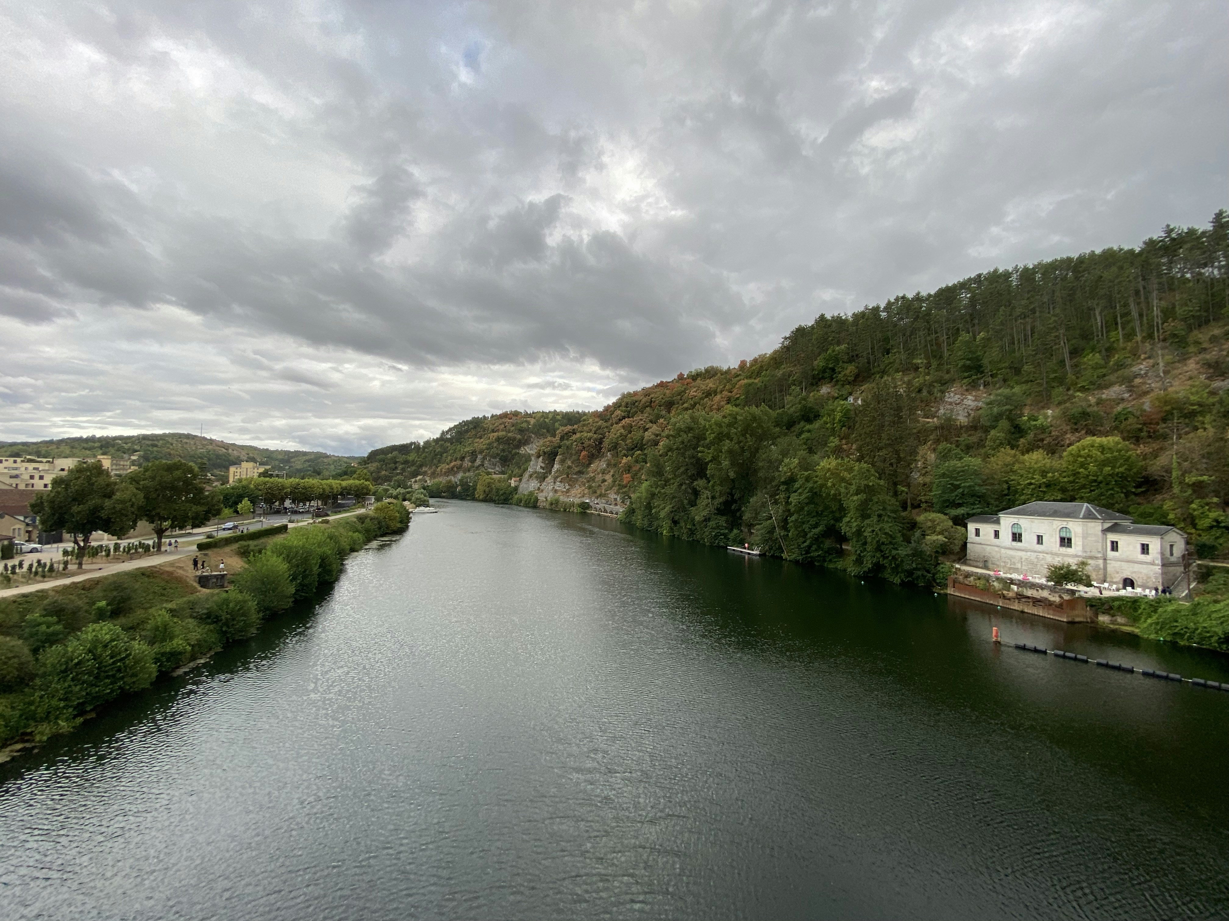 Wide river flanked by lush greenery and hills, with a house nestled on the riverbank beneath a cloudy sky.