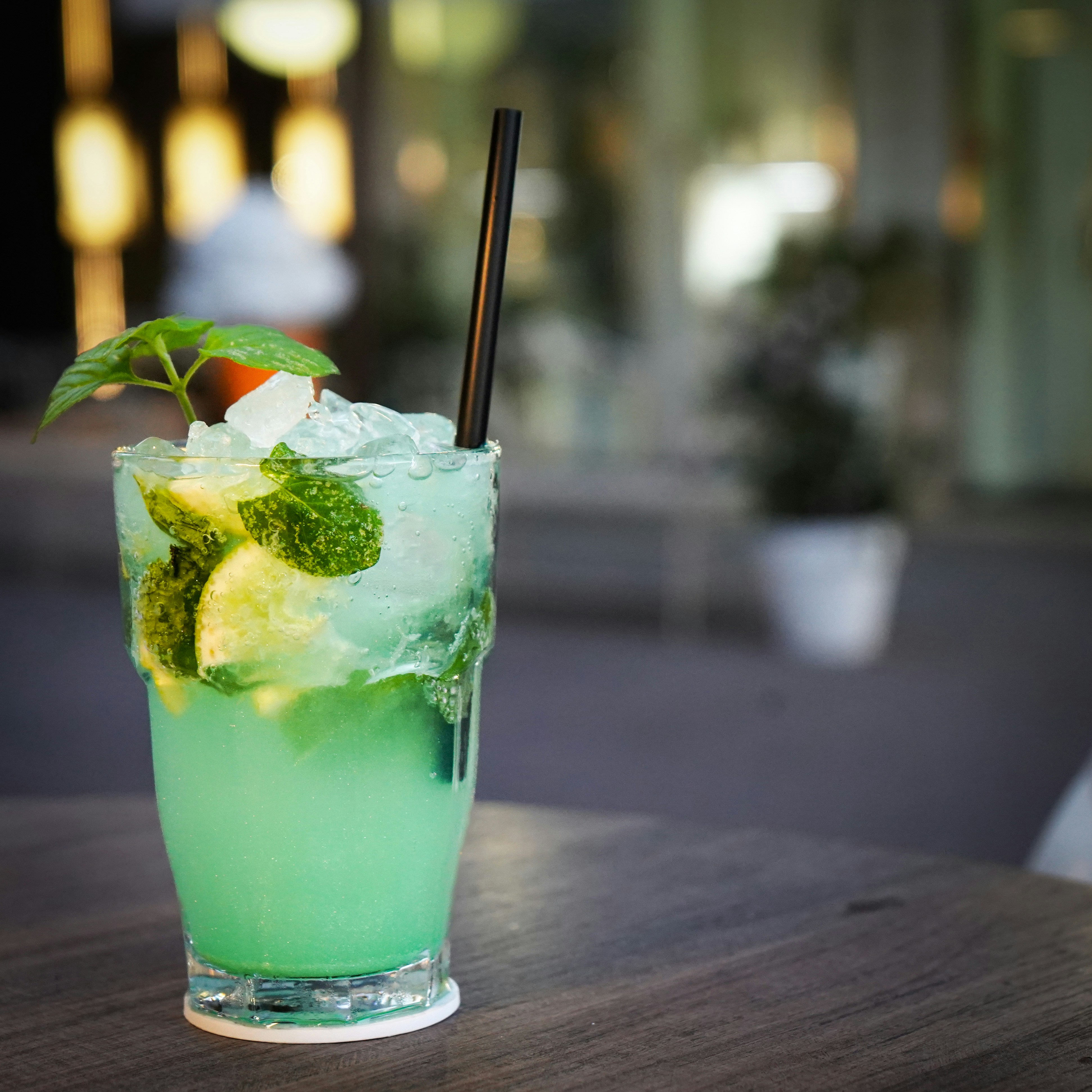 A vibrant cocktail garnished with mint and lime, served in a clear glass filled with ice. The background features softly blurred lights, enhancing the drink's appeal.