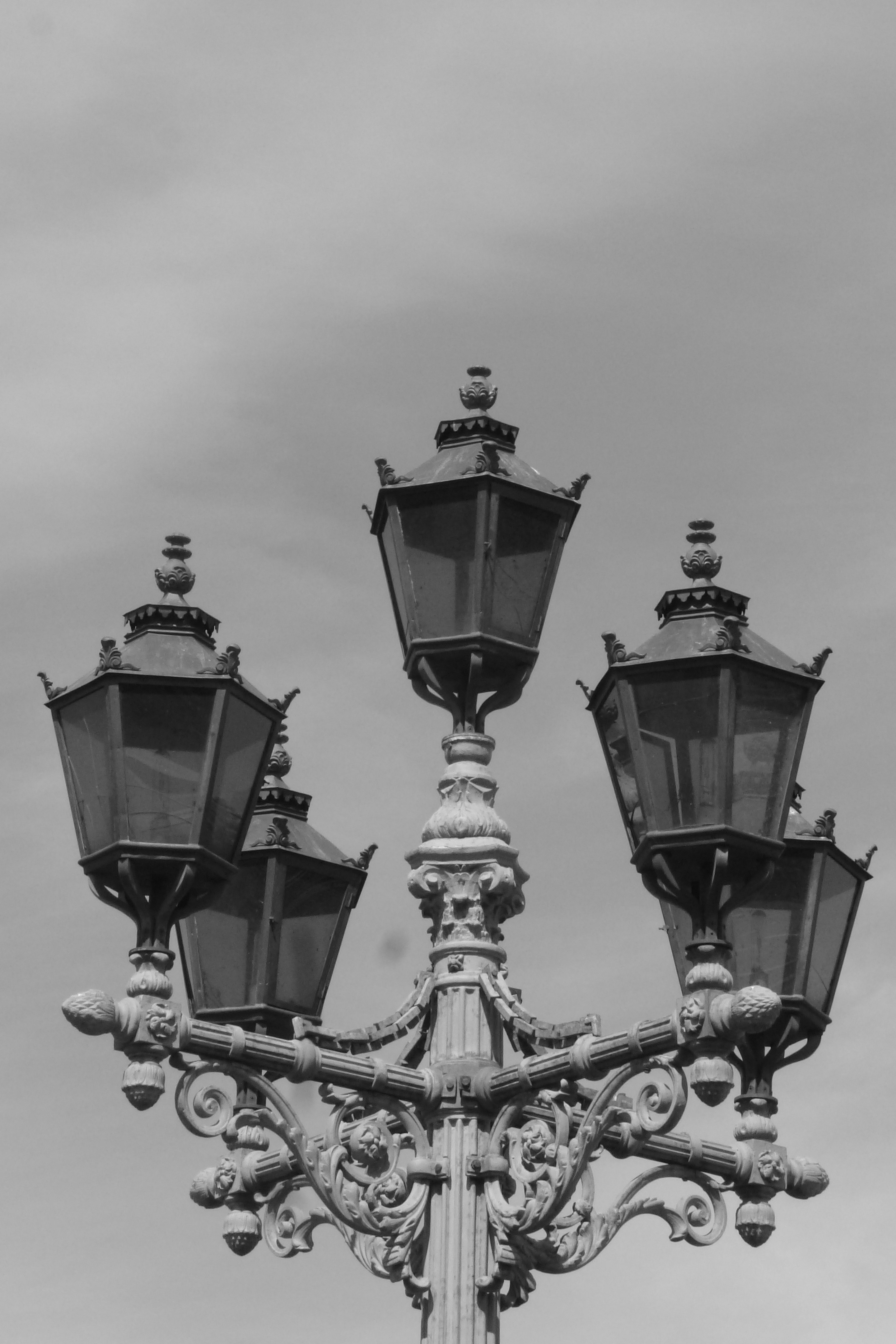 Black-and-white photograph of an ornate multi-lantern street lamp against a cloudy sky, highlighting intricate ironwork and symmetry.