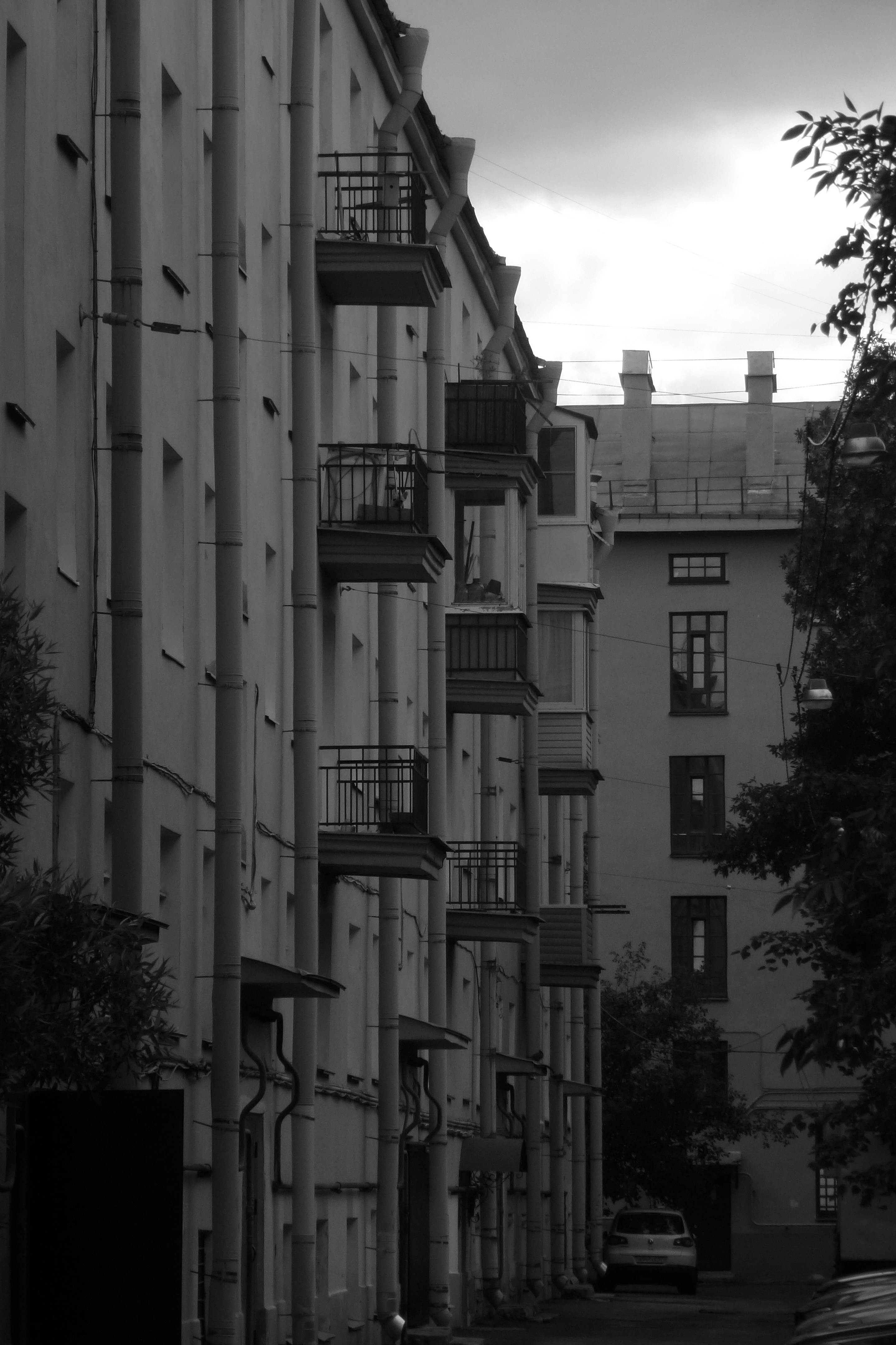 Black-and-white photograph of a row of apartment balconies along a narrow urban alley. A parked car and distant building anchor the scene.