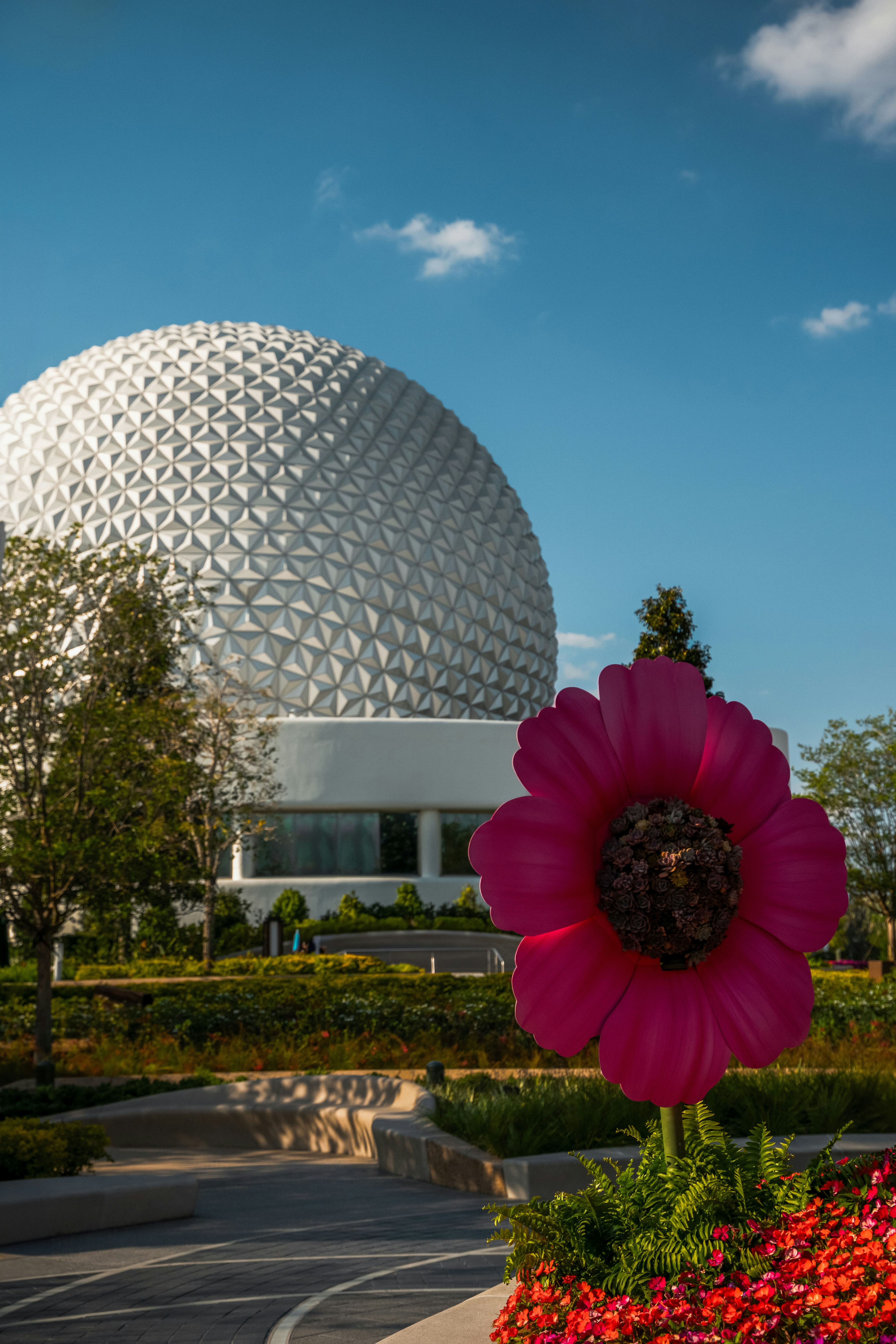 A bold magenta flower stands foreground with a geometric domed structure and blue sky in the background.