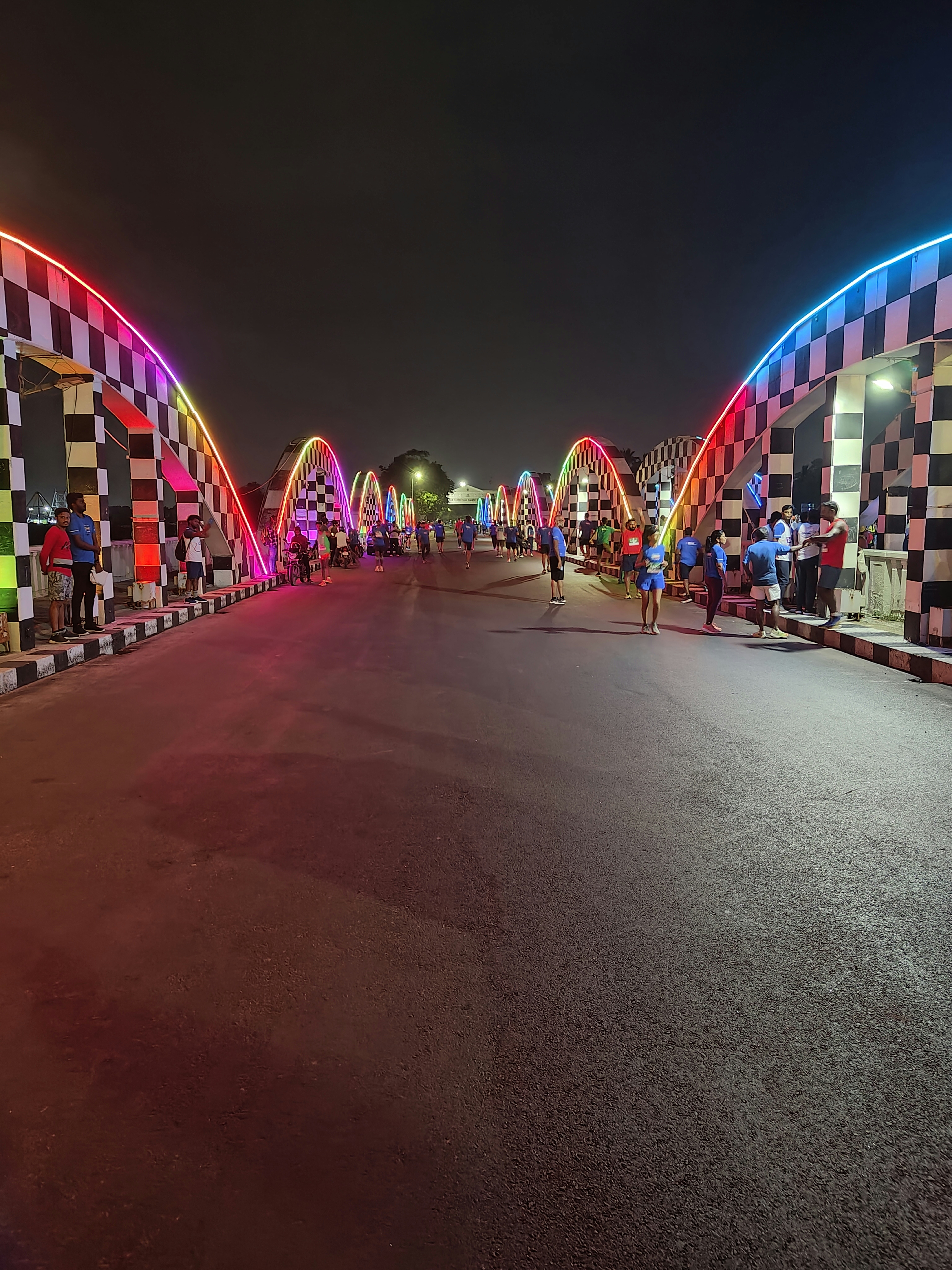 Night street photograph of neon rainbow arches lining a pedestrian promenade, with checker-pattern walls and people strolling.