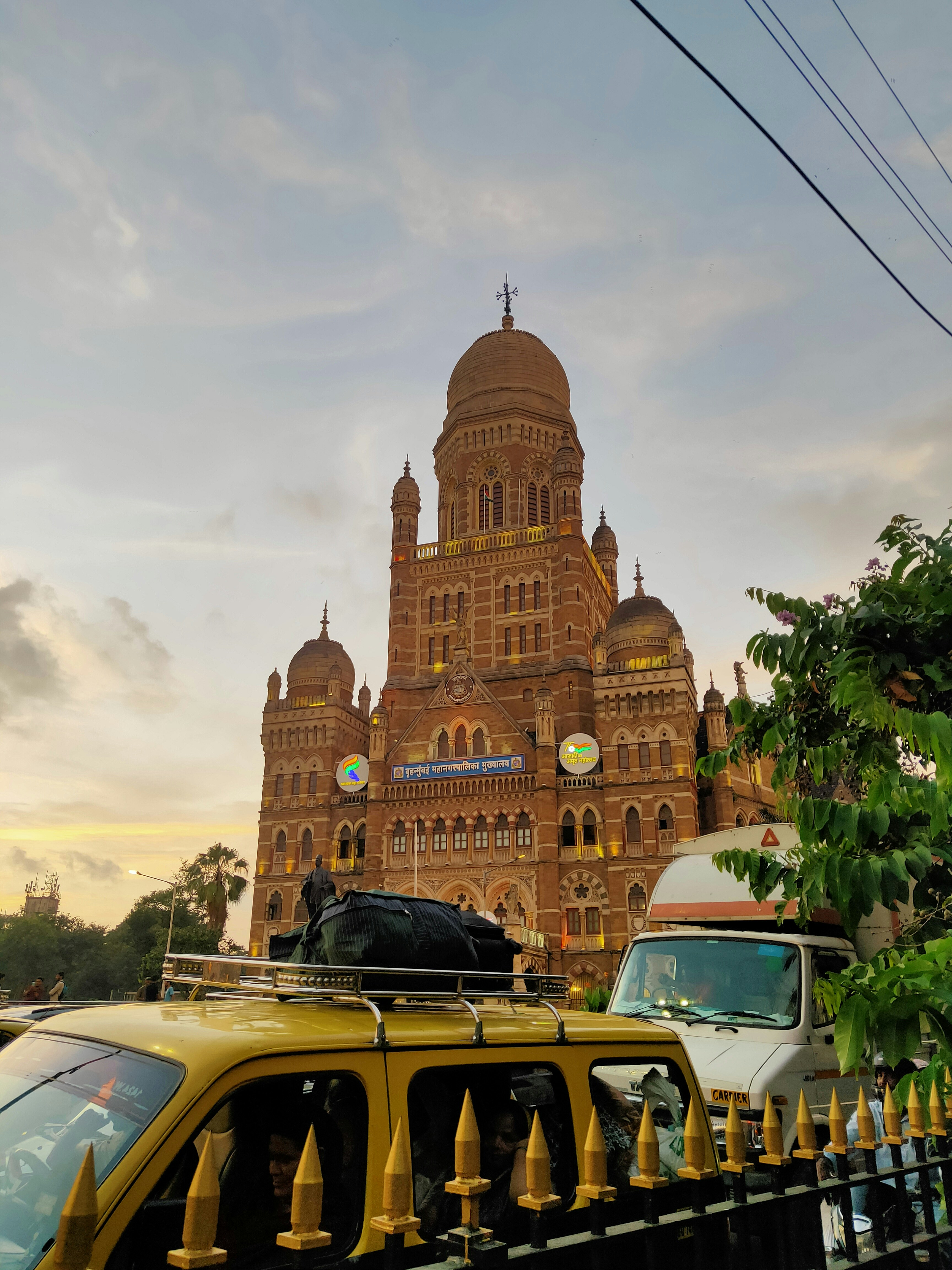 Photo of the Rajabai Clock Tower in Mumbai at golden hour, with yellow taxis and a white van in the foreground. The frame emphasizes the ornate brickwork and domed towers against a pastel sky.