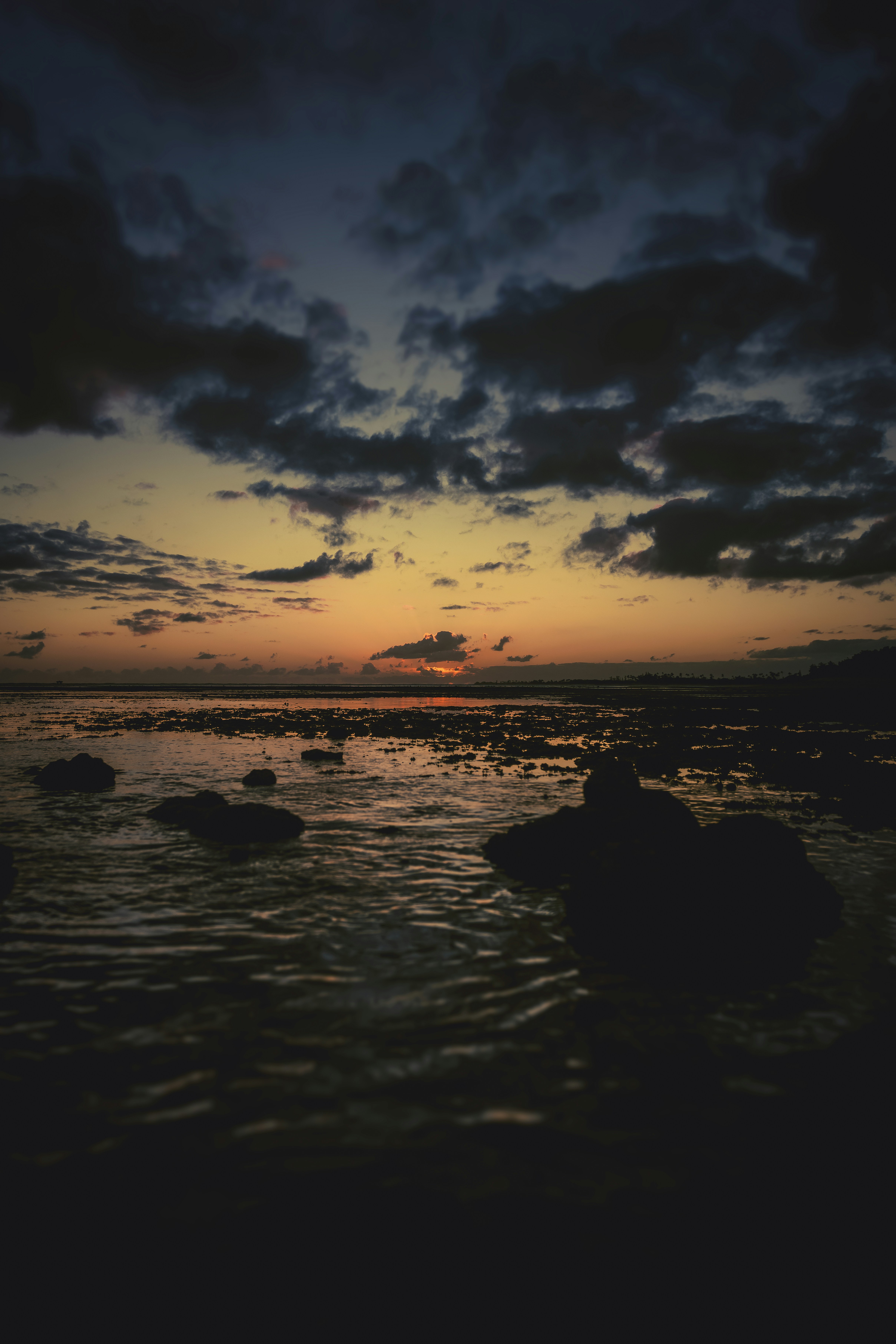 Dramatic clouds over a tranquil ocean at sunset, with rocks silhouetted against the colorful horizon.