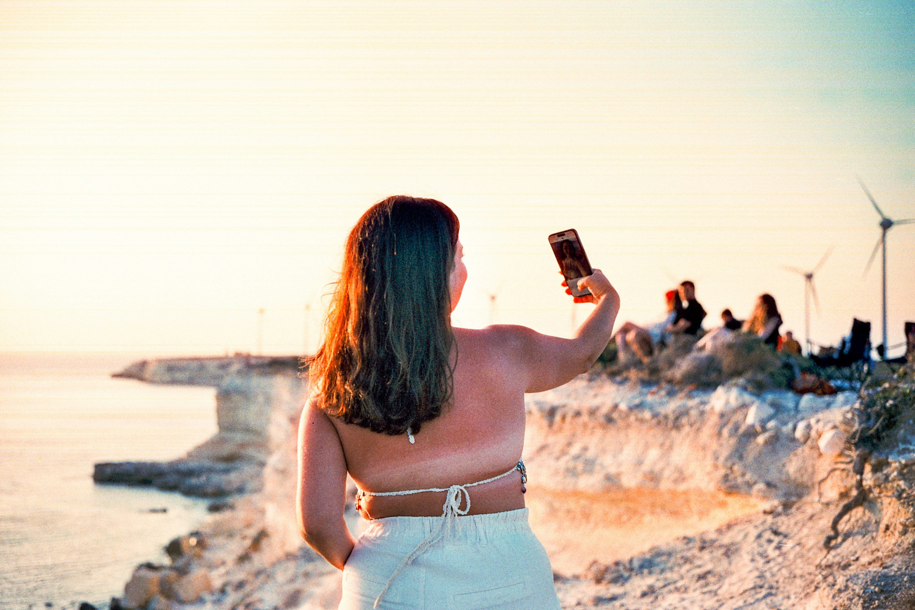 Person taking a selfie on a cliffside during sunset with wind turbines in the background.