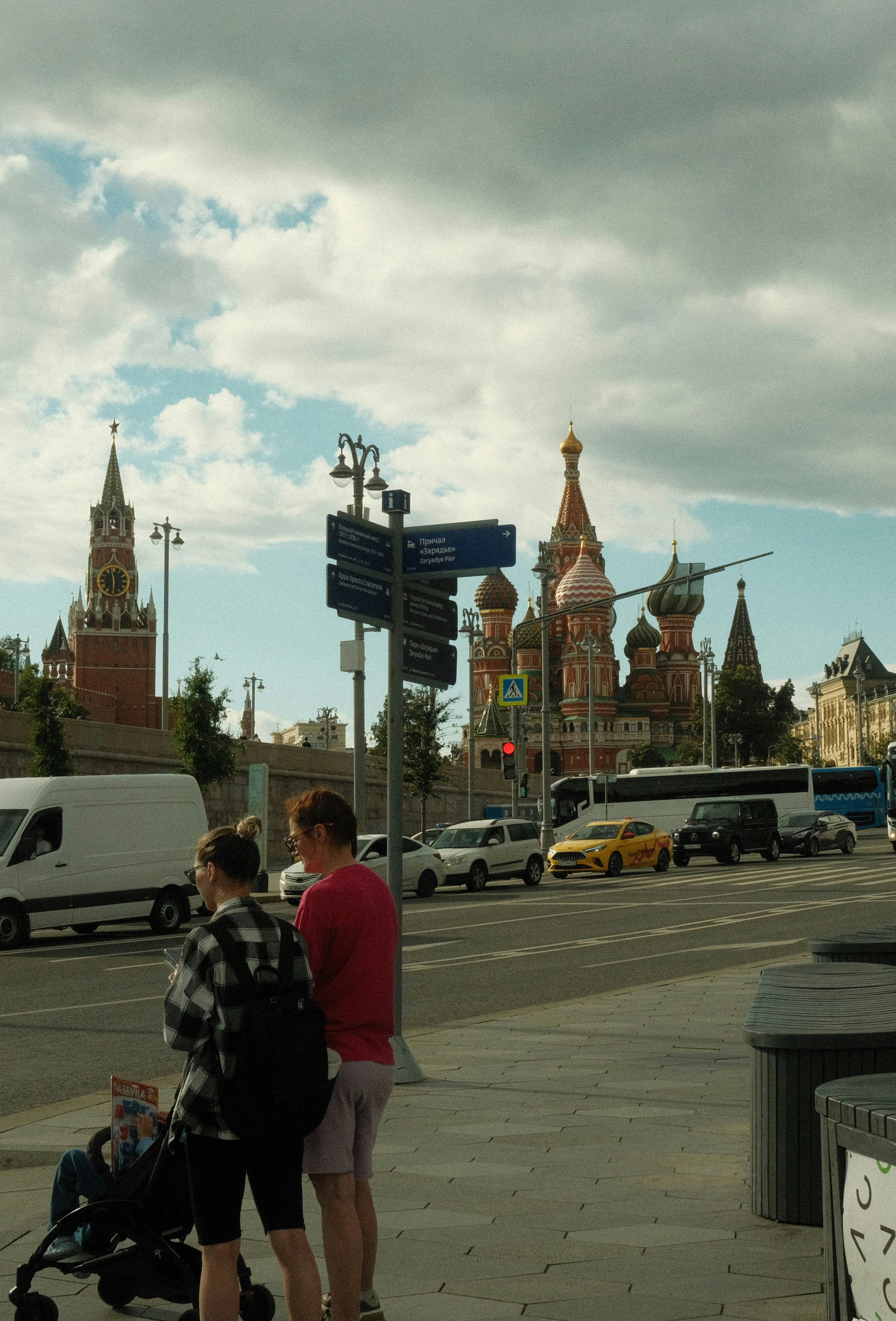 A street scene in Moscow with St. Basil's Cathedral in the background as a couple with a stroller waits near a crosswalk while traffic rolls by.