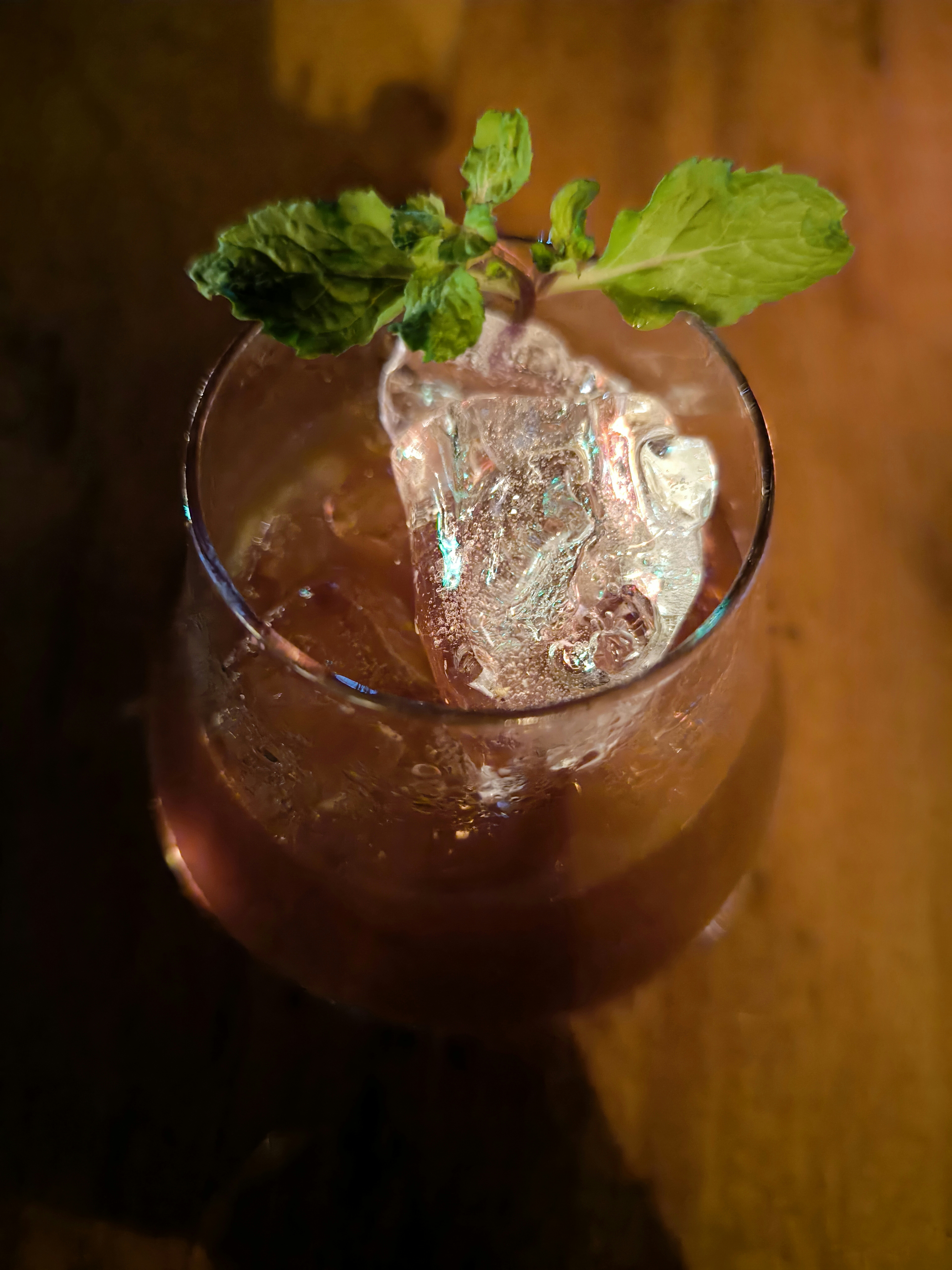 A cocktail glass filled with ice and a mint garnish sits on a wooden surface. Warm lighting highlights the glass contours and the mint's fresh texture.