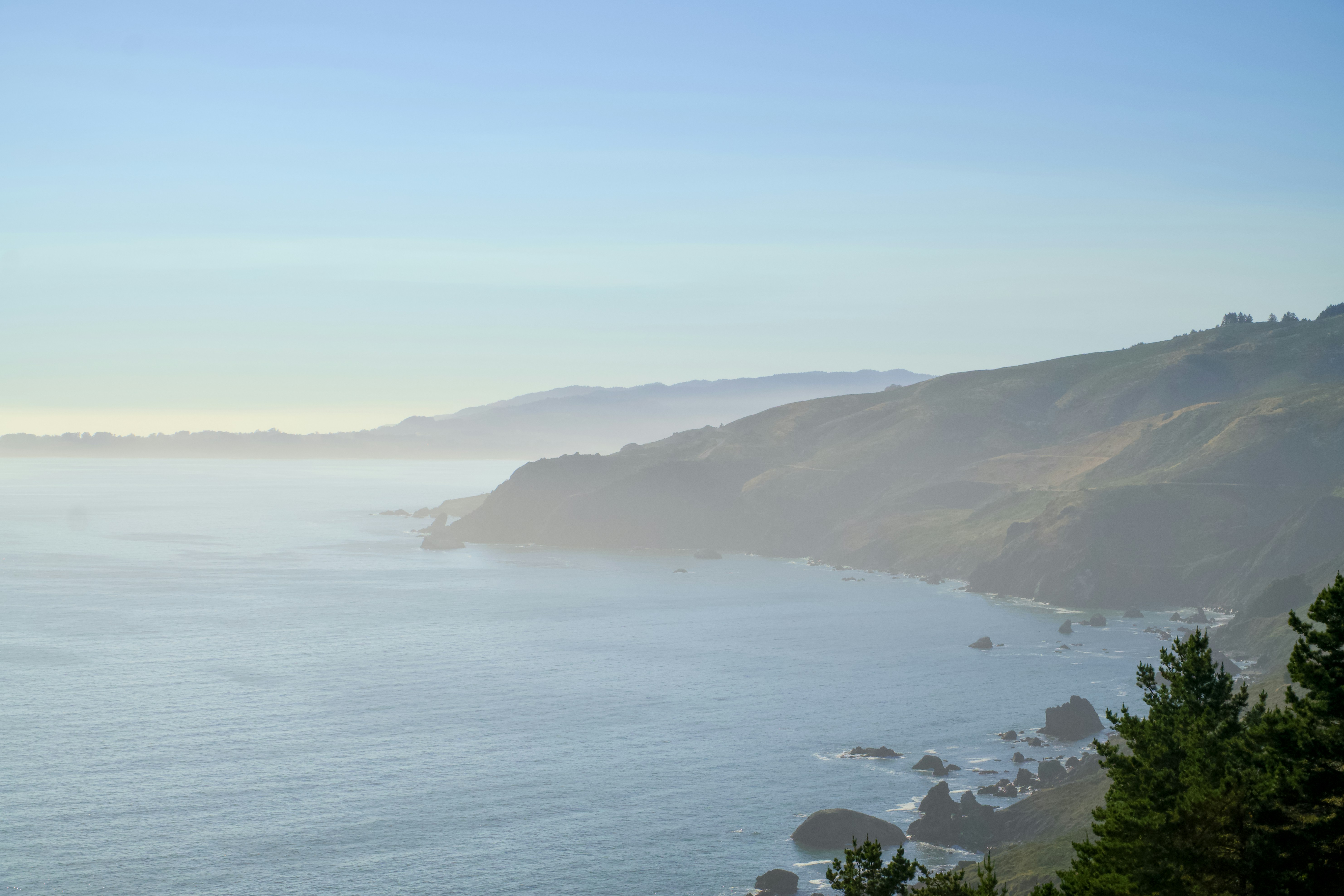 Gentle waves lap against a misty coastline, where rolling hills meet the ocean under a serene sky.