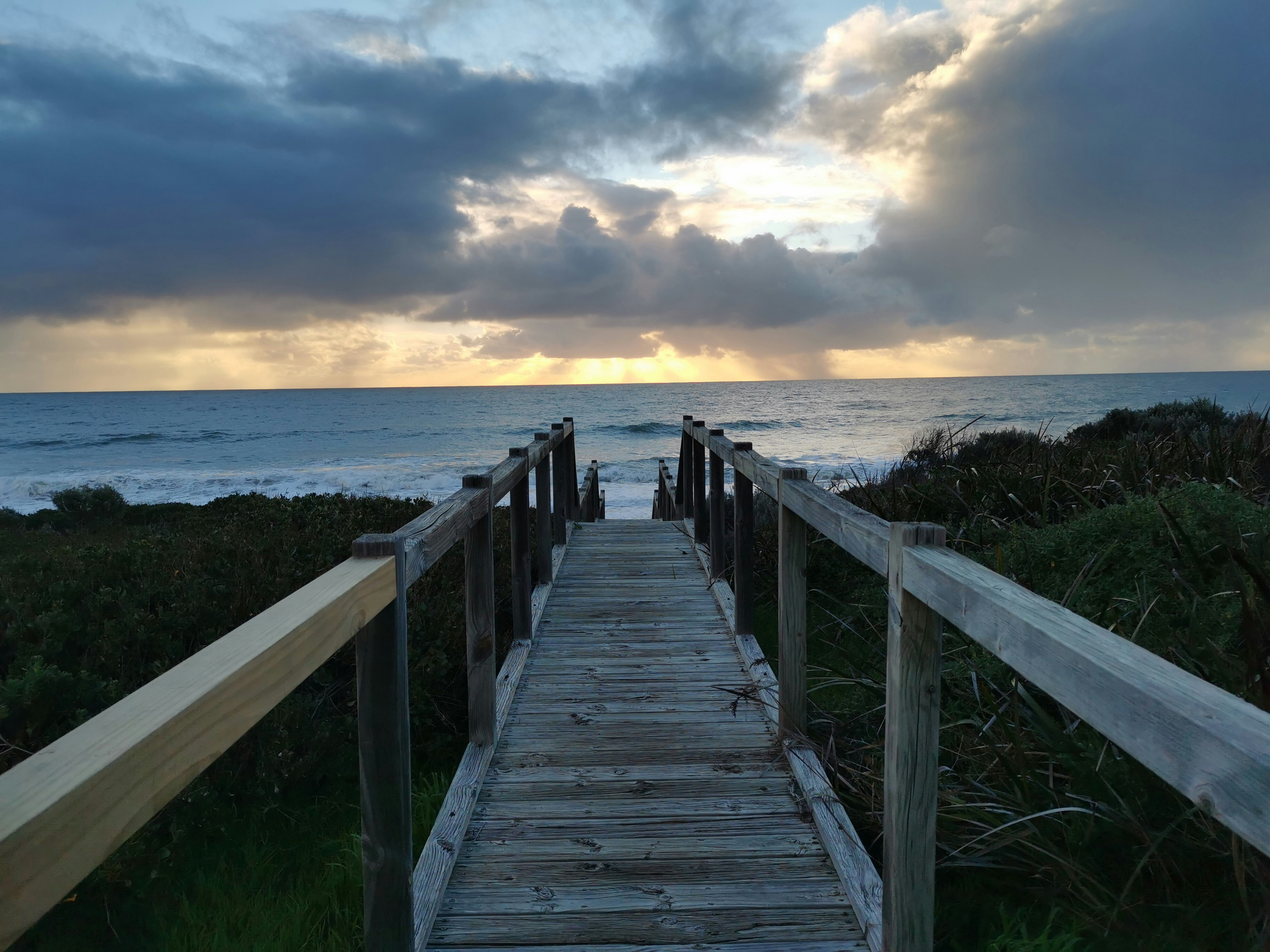 Wooden walkway leading to a vast ocean horizon beneath a dramatic cloudy sky.