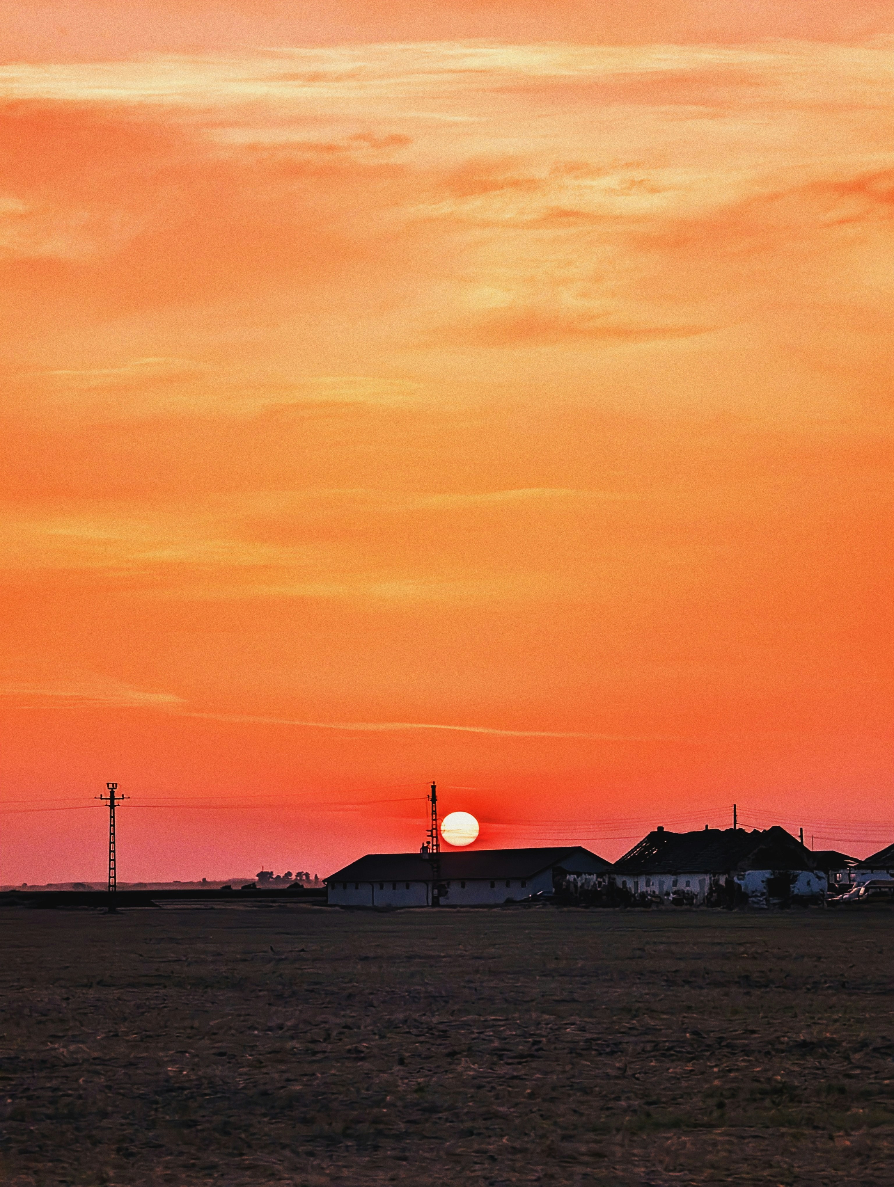 Vibrant sunset casting warm hues over silhouetted buildings and power lines on an open landscape. The sun dips below the horizon, creating a tranquil atmosphere.