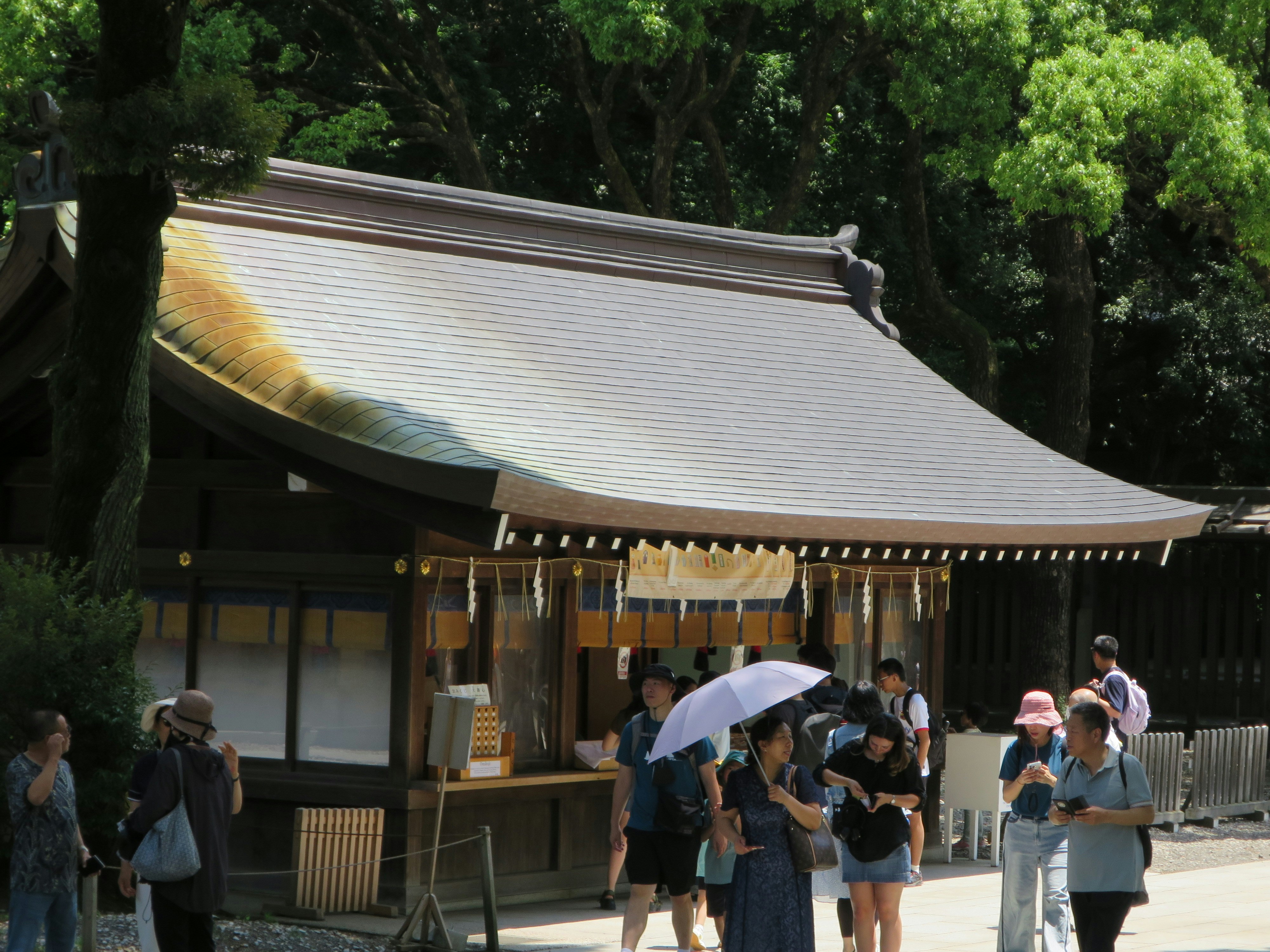 Visitors gather in a Shinto shrine courtyard as a curved roof and shimenawa frame the scene, with a person holding a white umbrella among the group.