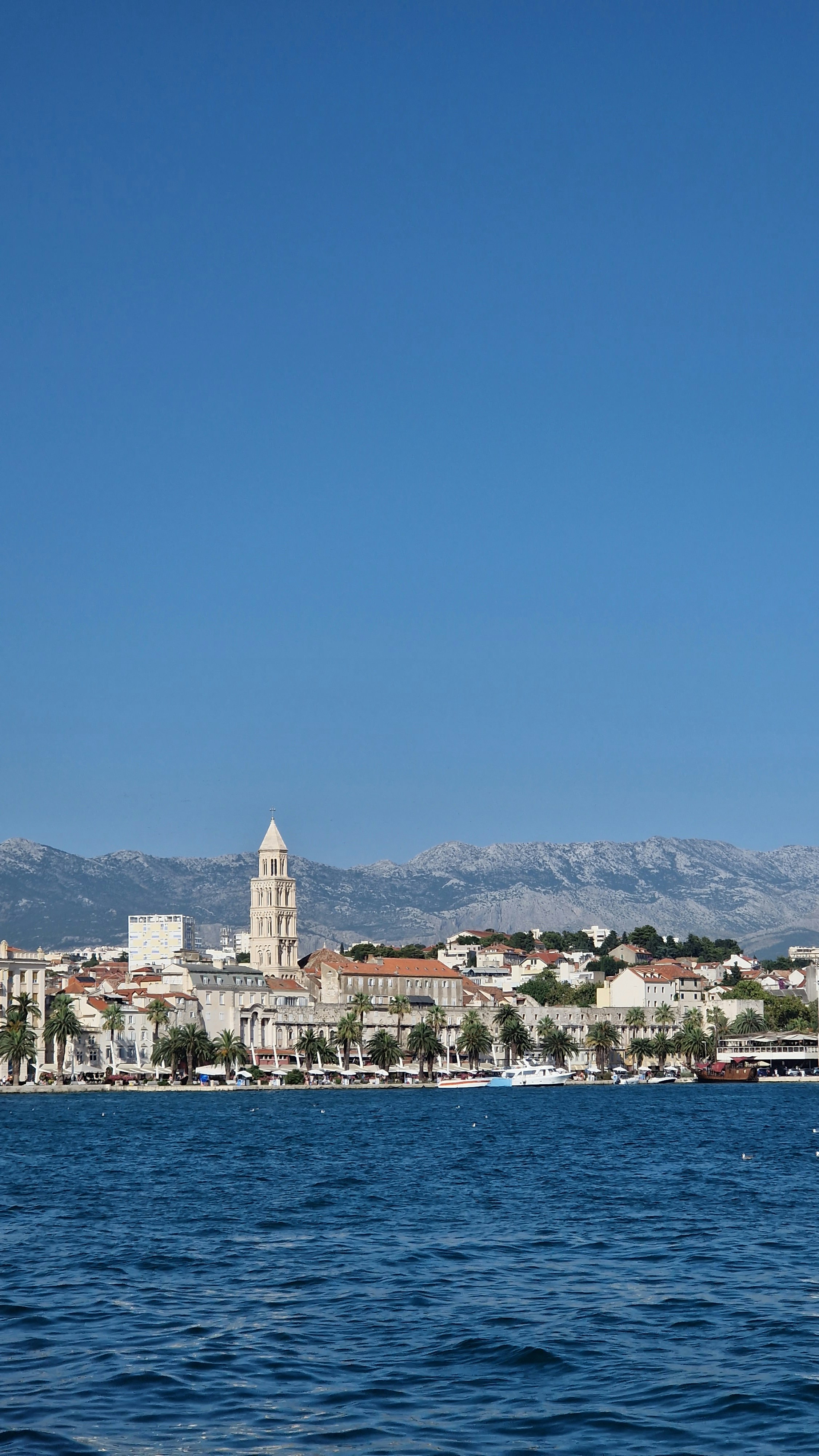 Photograph of a coastal town with a palm-lined promenade, a tall clock tower, red-tiled buildings along the shore, and a calm blue sea under a clear sky.