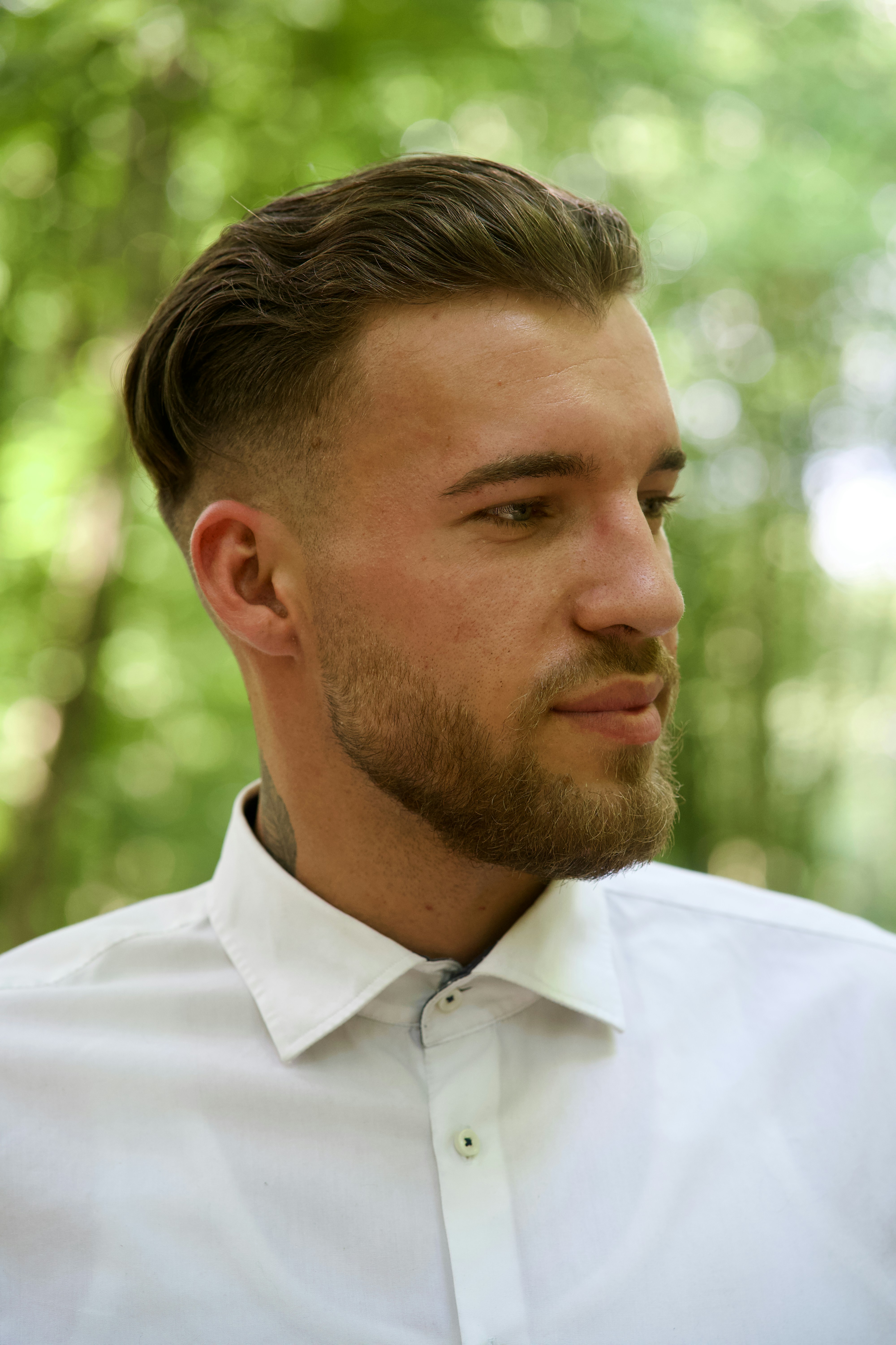 Man in a white shirt and tie gazing thoughtfully with a blurred green forest backdrop.