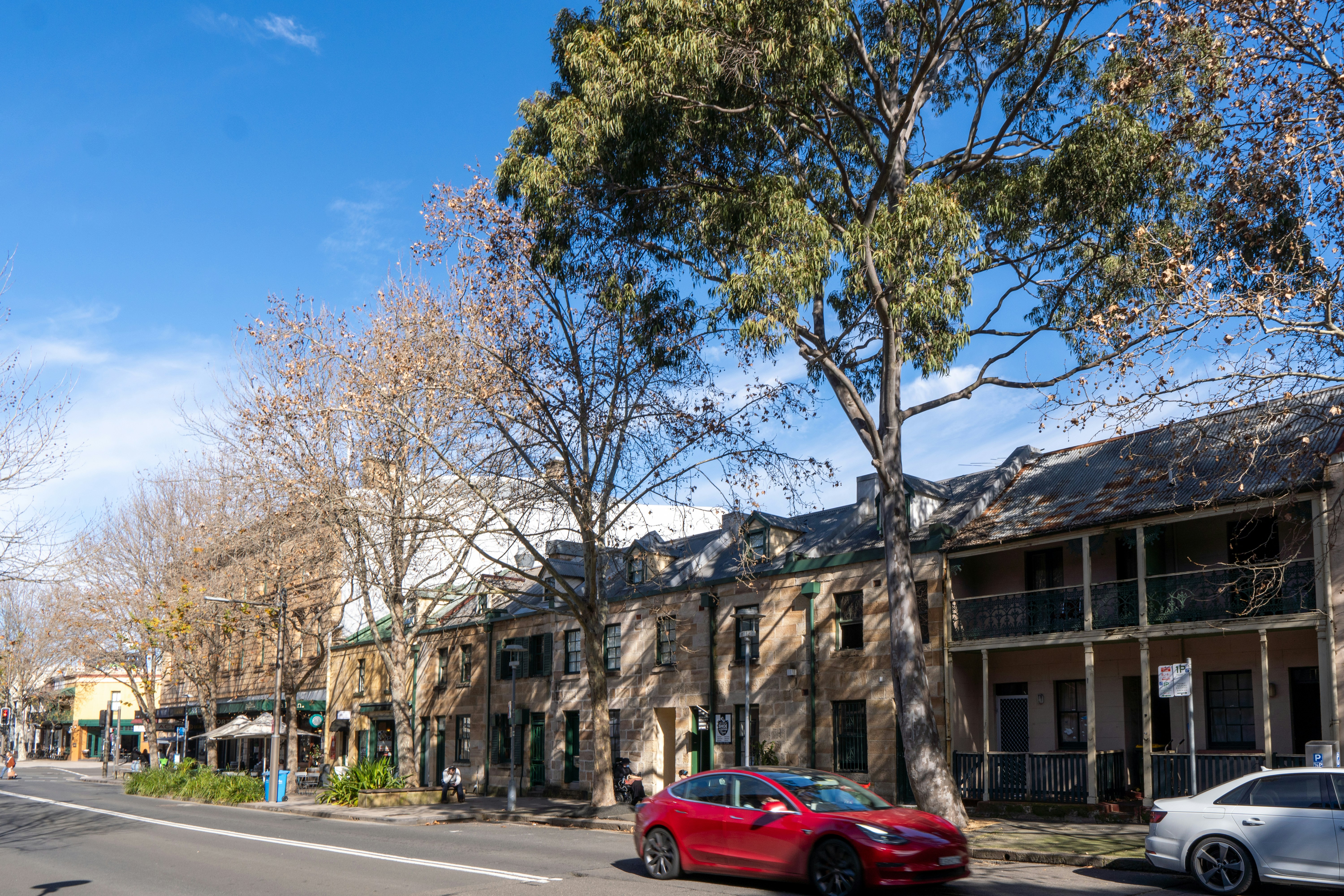 Historic buildings line a peaceful street, framed by towering trees and a clear blue sky. A red car adds a splash of color to the tranquil scene.