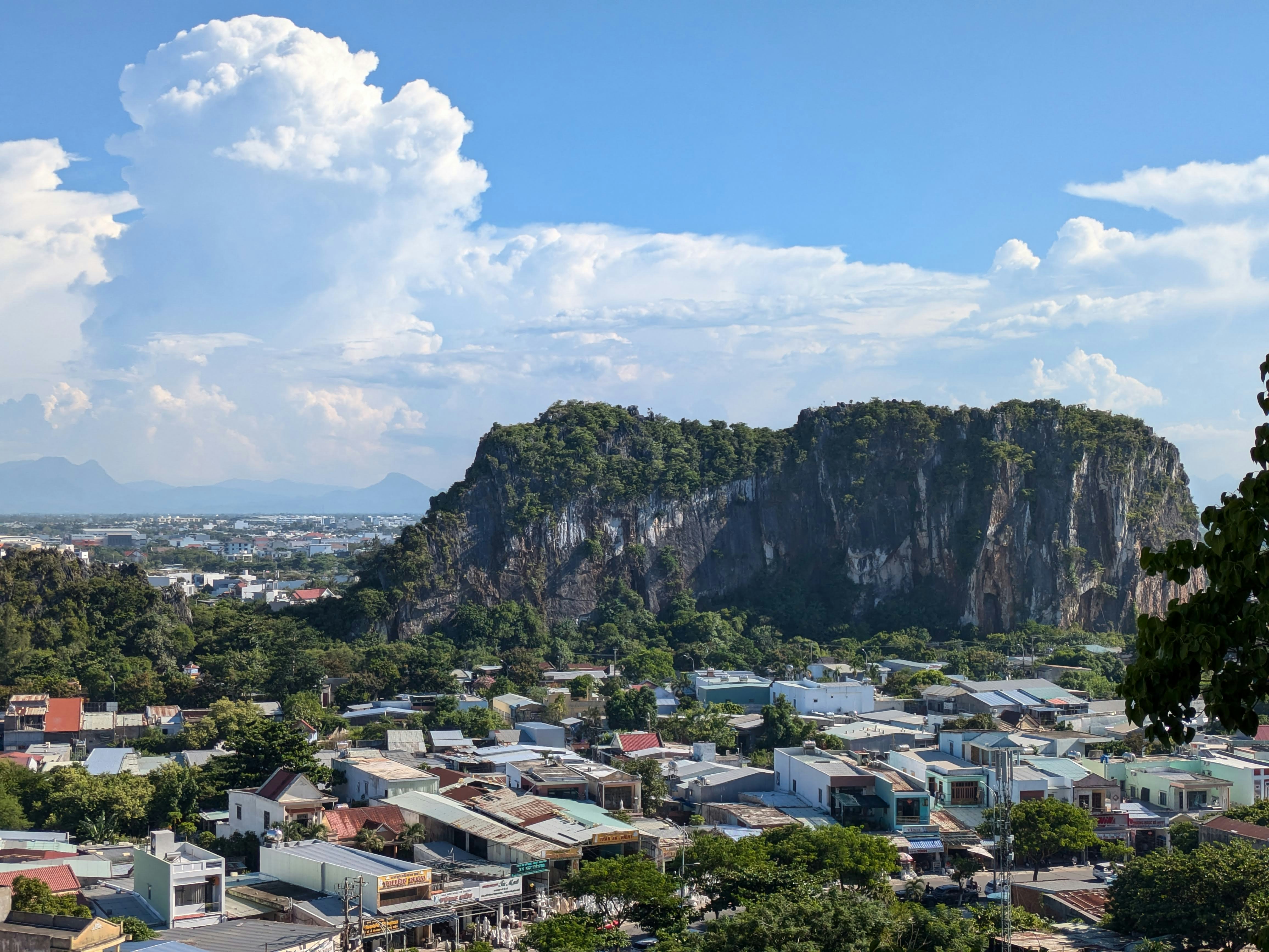 A panoramic view showcasing a rugged limestone formation amidst a vibrant urban landscape, under a bright blue sky with scattered clouds.