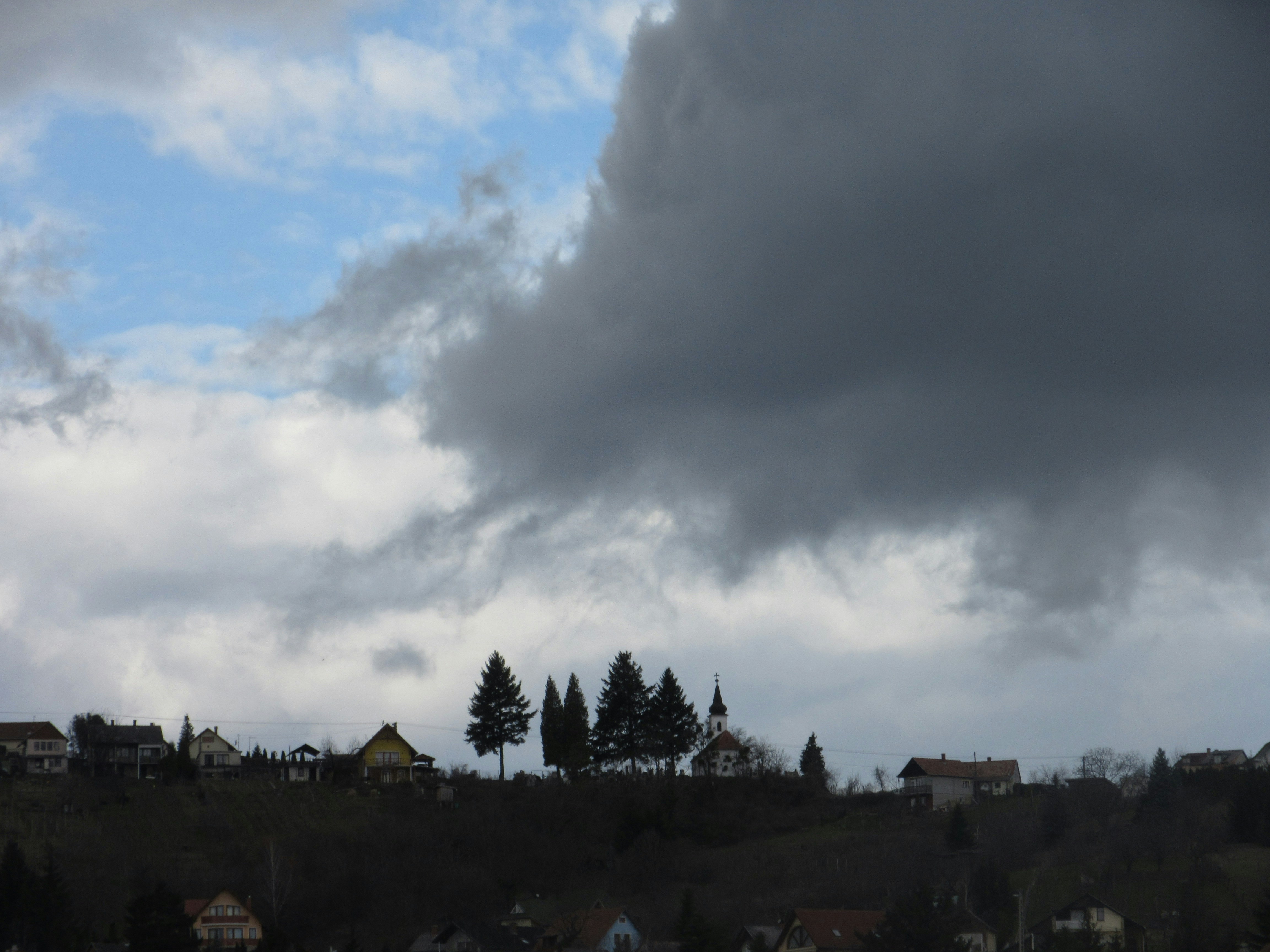 Horizon line with a row of houses and a cluster of trees on a gentle hillside under a dramatic, cloud-filled sky.
