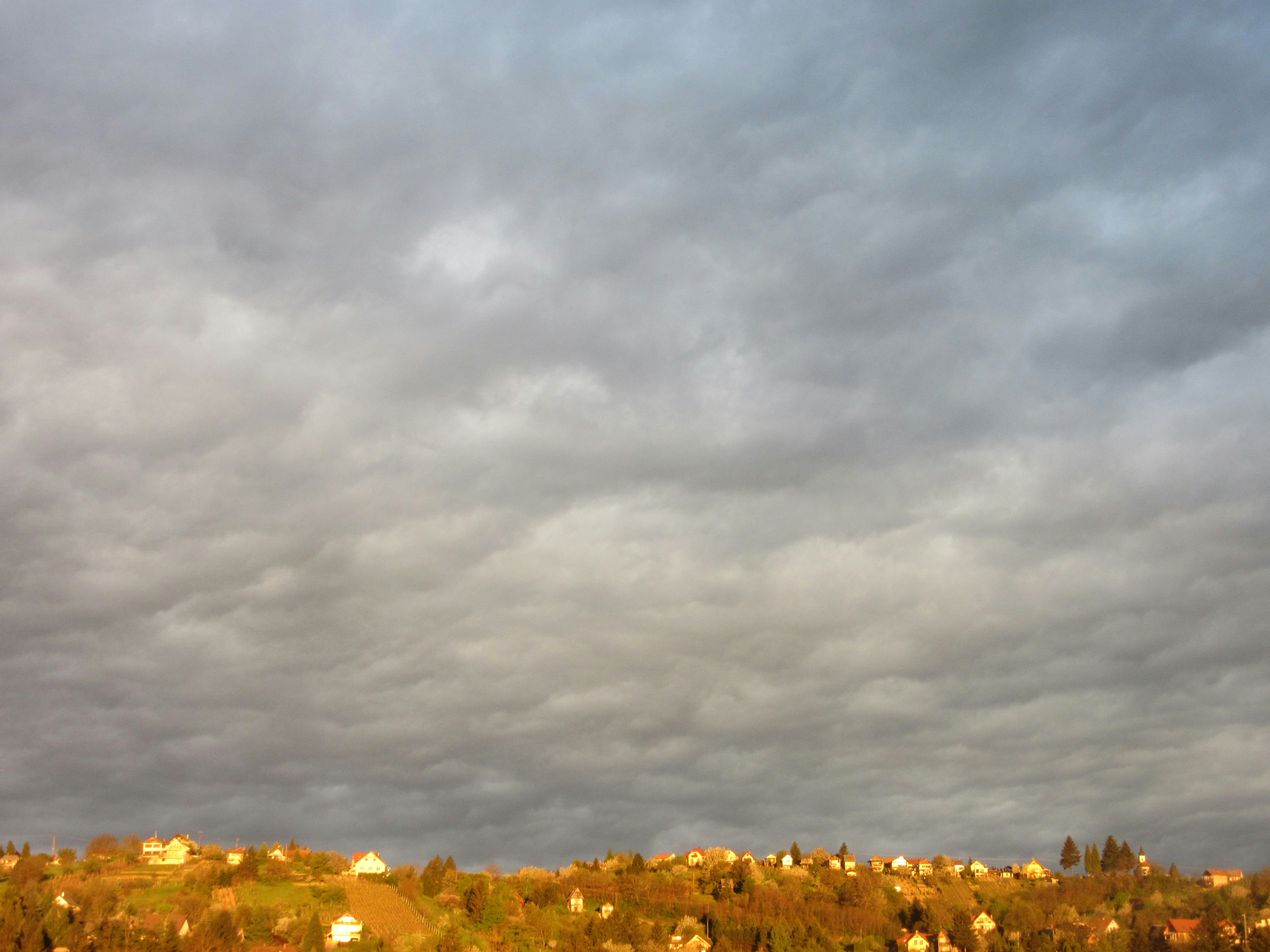 Row of hillside houses bathed in warm light along a low horizon, beneath a dramatic, cloud-filled sky.
